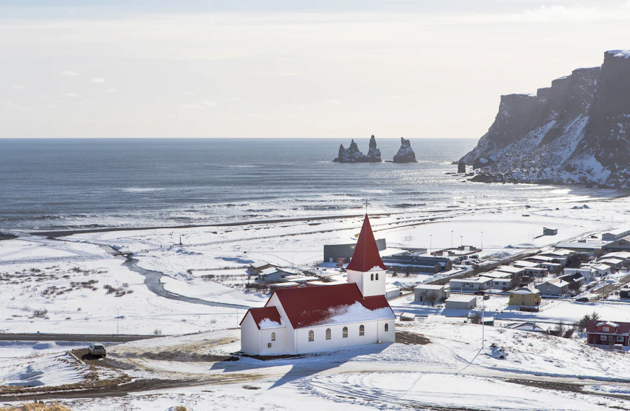 White church with a red roof and steeple sits on a snowy seaside village, with dark cliffs and sea stacks beyond the frozen shoreline.