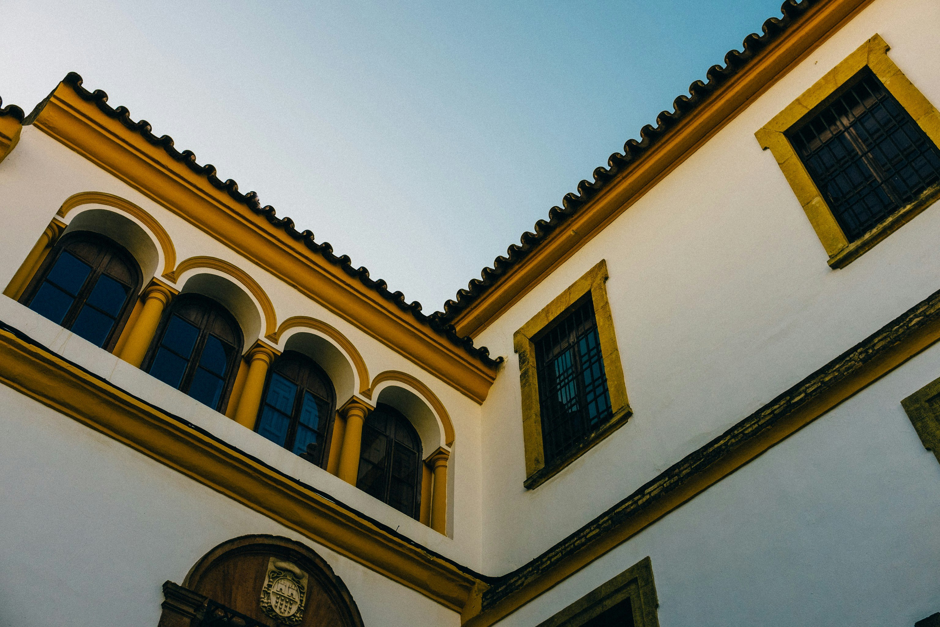 Upward view of a Mediterranean-style building facade with arched windows and contrasting yellow trim against a clear blue sky.
