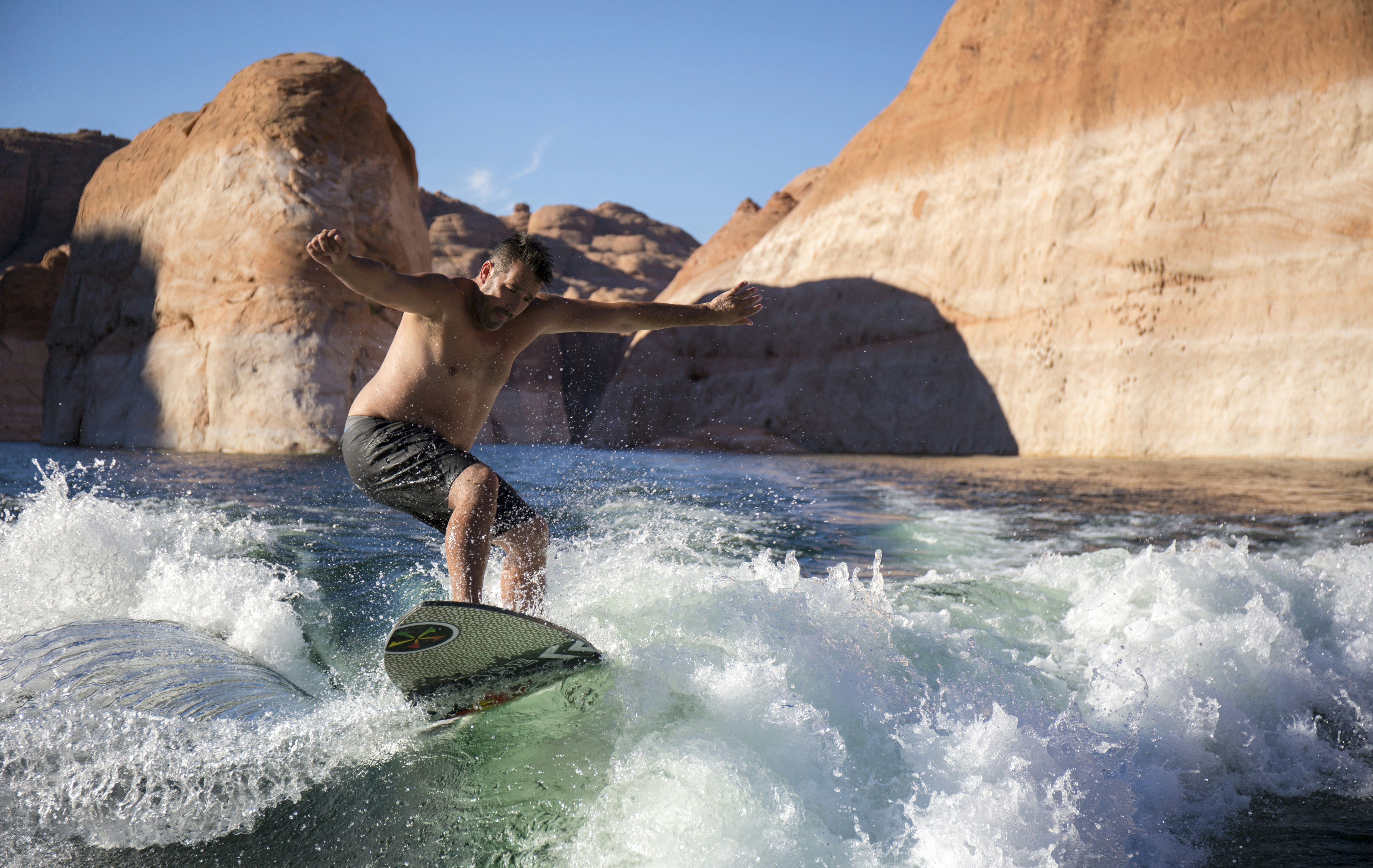 Surfer riding a wave against a backdrop of towering red rock formations under a clear blue sky.