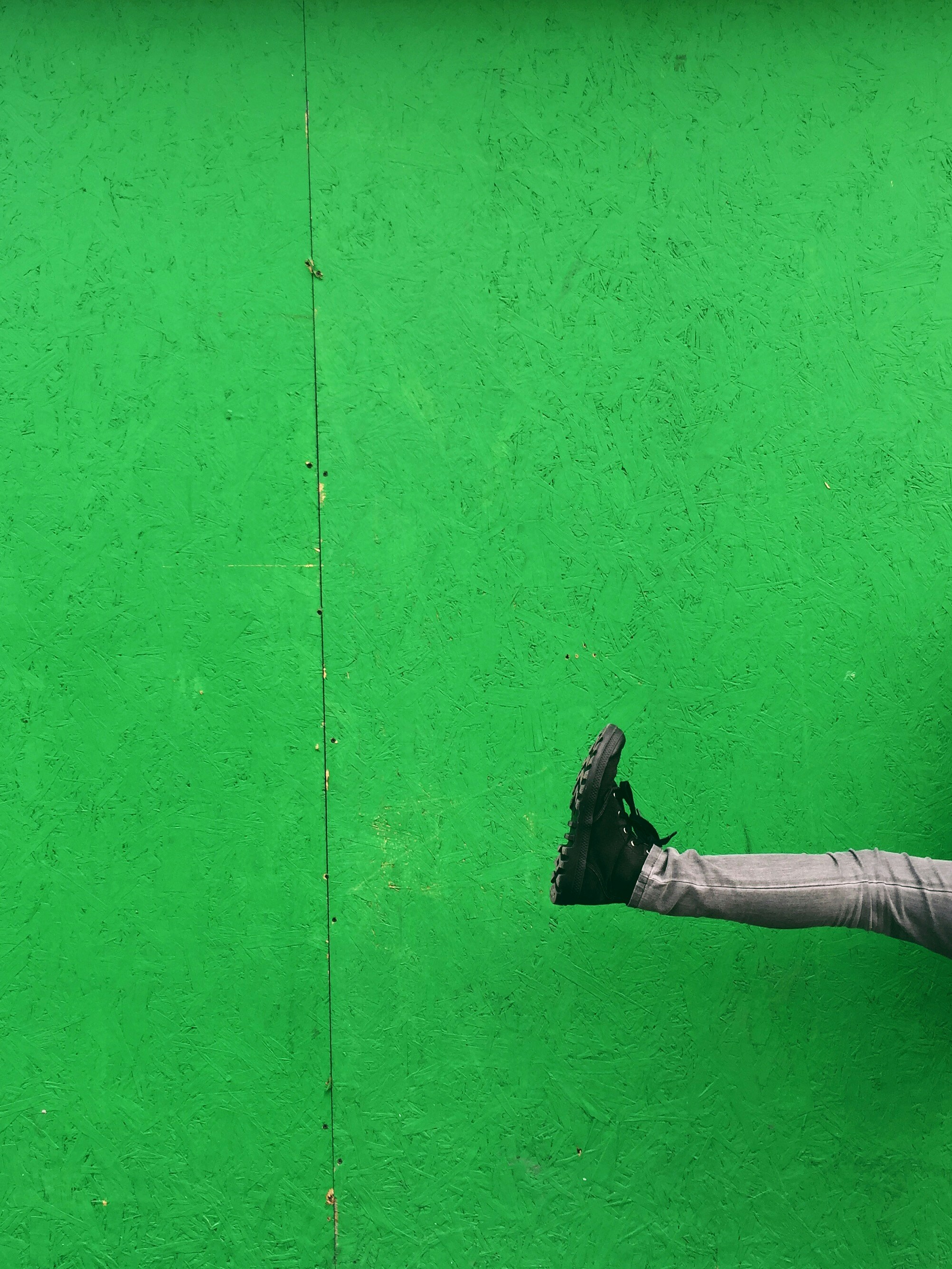 A foot clad in a black boot is raised against a vibrant green wall, highlighting a playful urban moment.