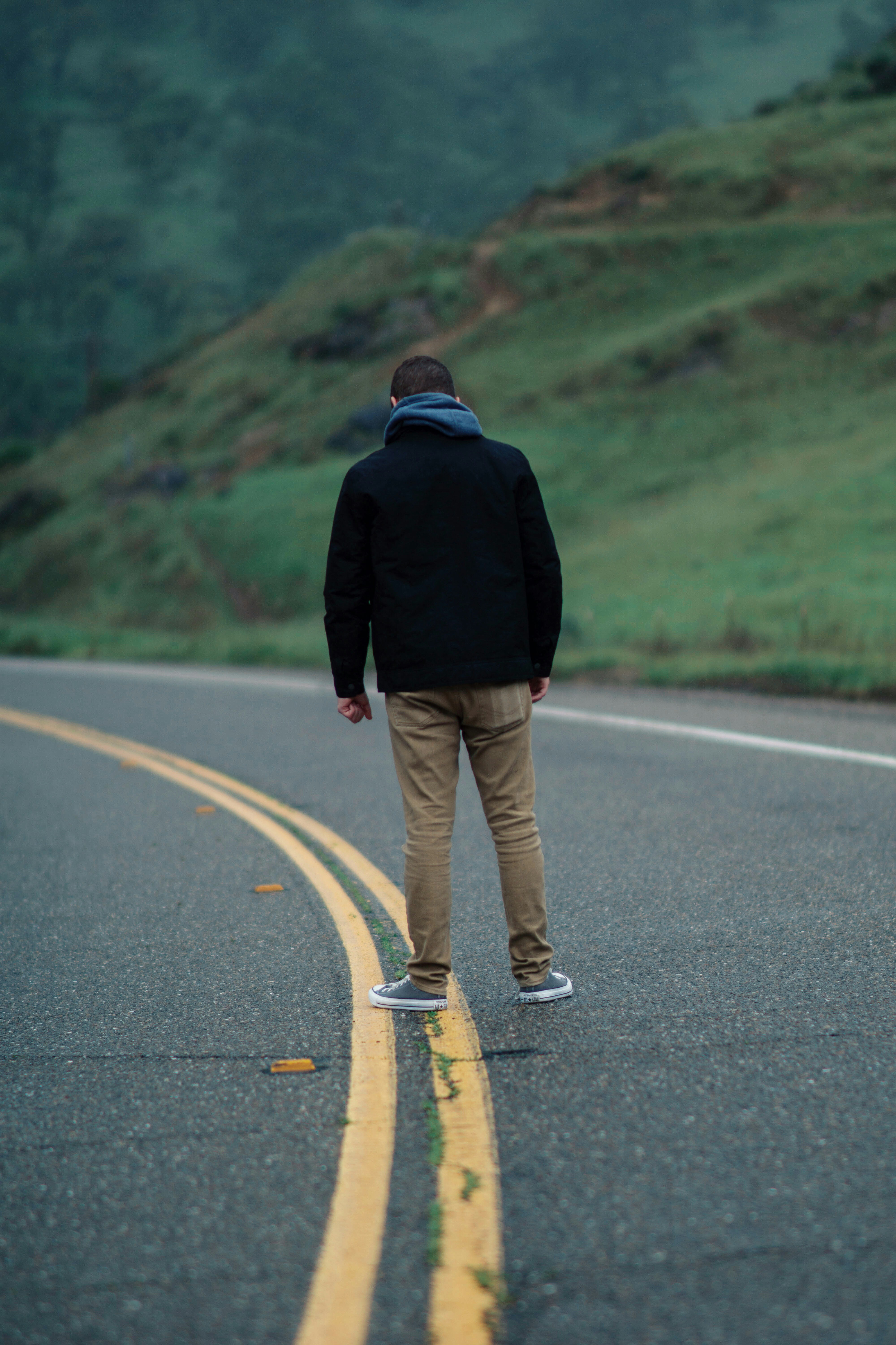 A figure stands on a winding road, surrounded by lush green hills under a moody sky. The composition emphasizes solitude and contemplation.