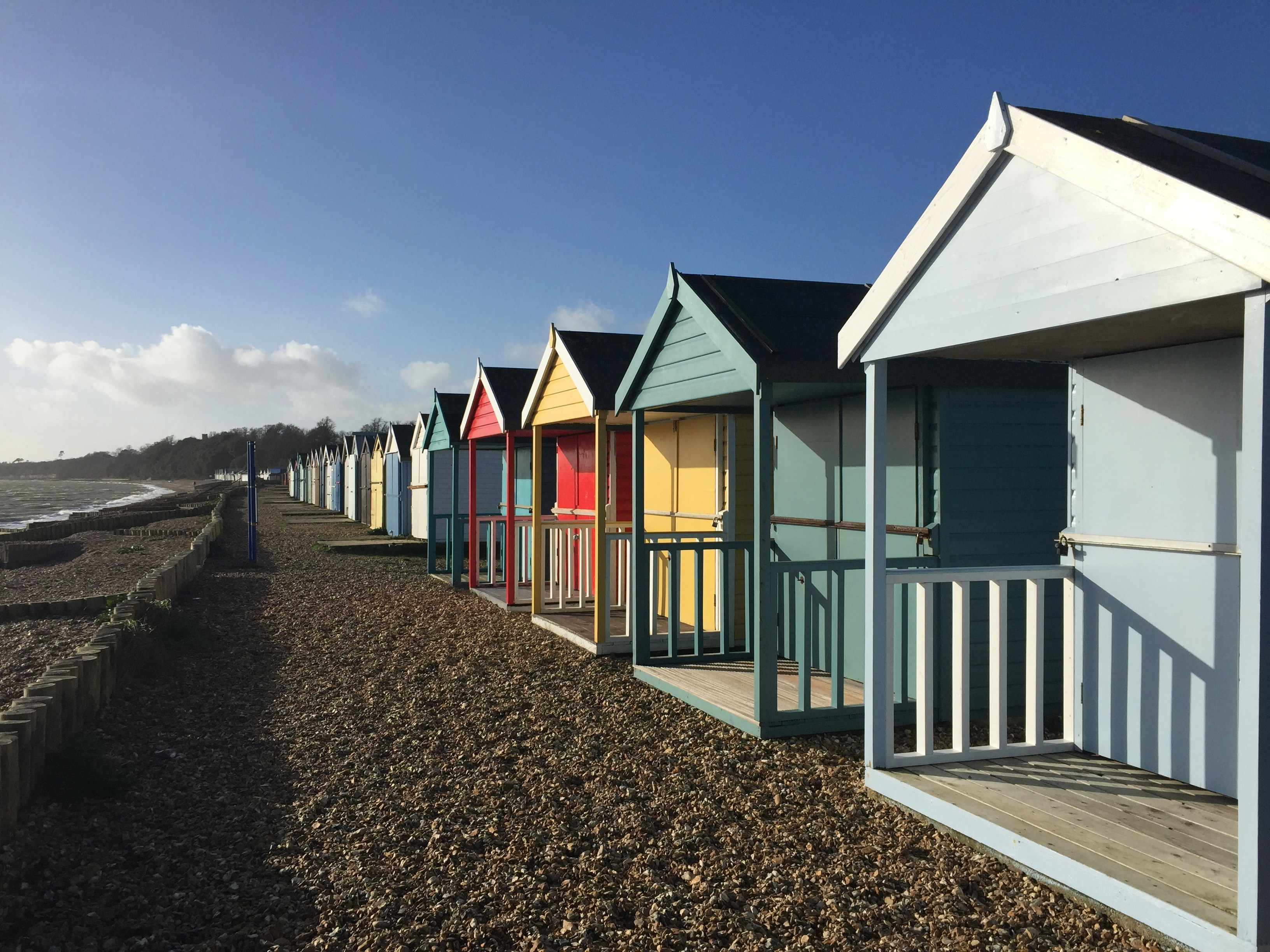 Colorful beach huts line a pebble shore under a clear blue sky.