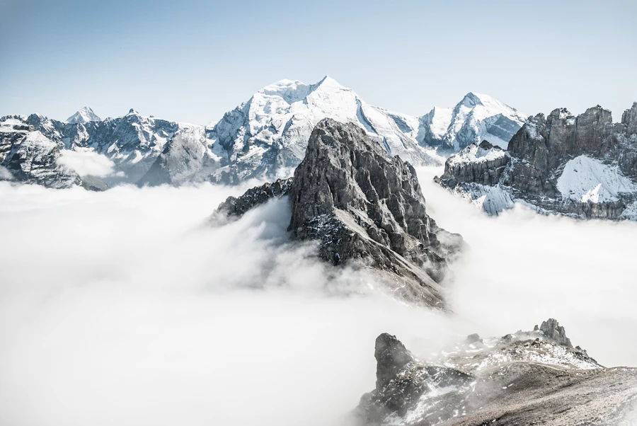 Jagged snow-covered mountain peaks rise above a blanket of clouds, with a dark central spire dominating the scene. Pale blue sky and misty foreground create a cold alpine atmosphere.