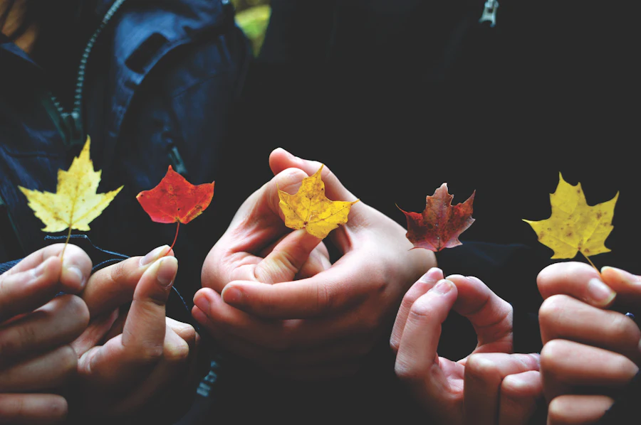 Several hands hold colorful autumn leaves in a close-up, forming a cooperative circle that centers on a yellow leaf. A soft, dark background emphasizes the leaves and fingertips; conveys theme of unity and season transition.