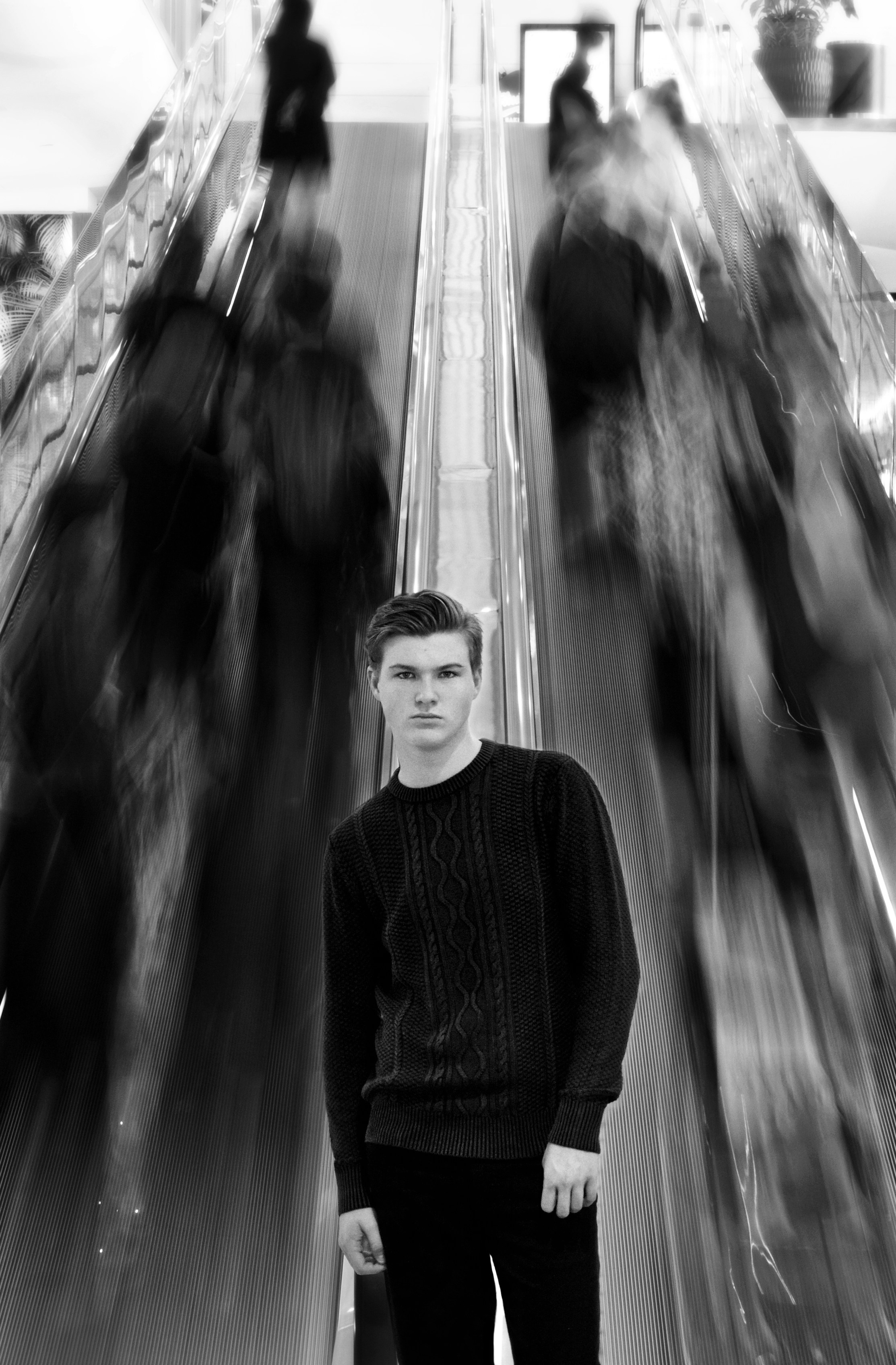 A young man stands confidently on an escalator, surrounded by a blur of hurried pedestrians in motion. The monochrome tones enhance the contrast between stillness and chaos.