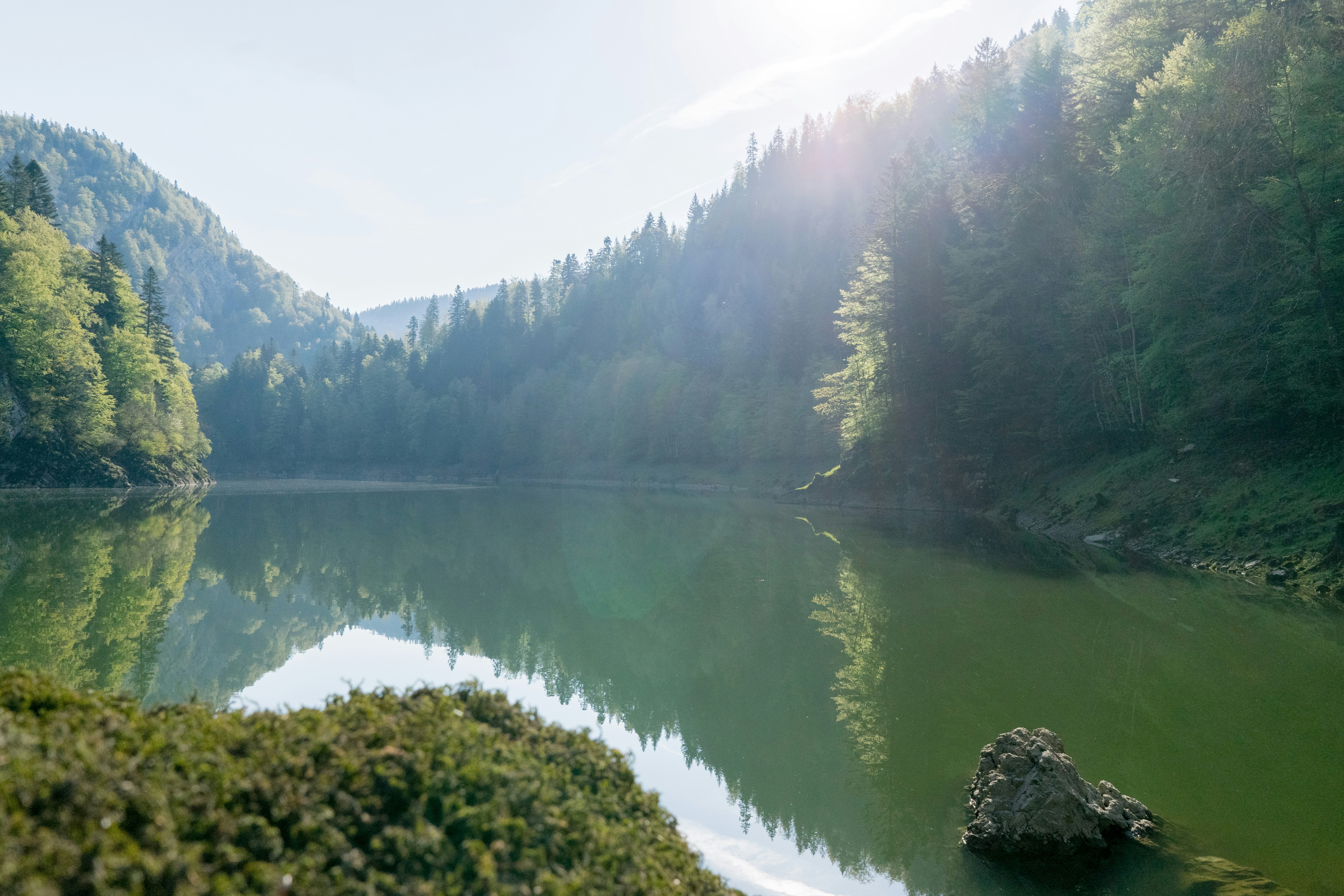 Tranquil lake reflecting lush green hills under soft sunlight, with moss-covered rocks in the foreground. A peaceful natural scene invites contemplation.