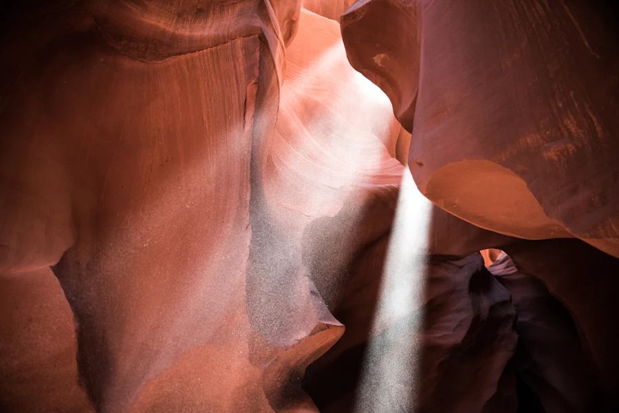 Photograph of a narrow slot canyon with a single beam of sunlight piercing warm red sandstone, revealing smooth, sculpted walls.