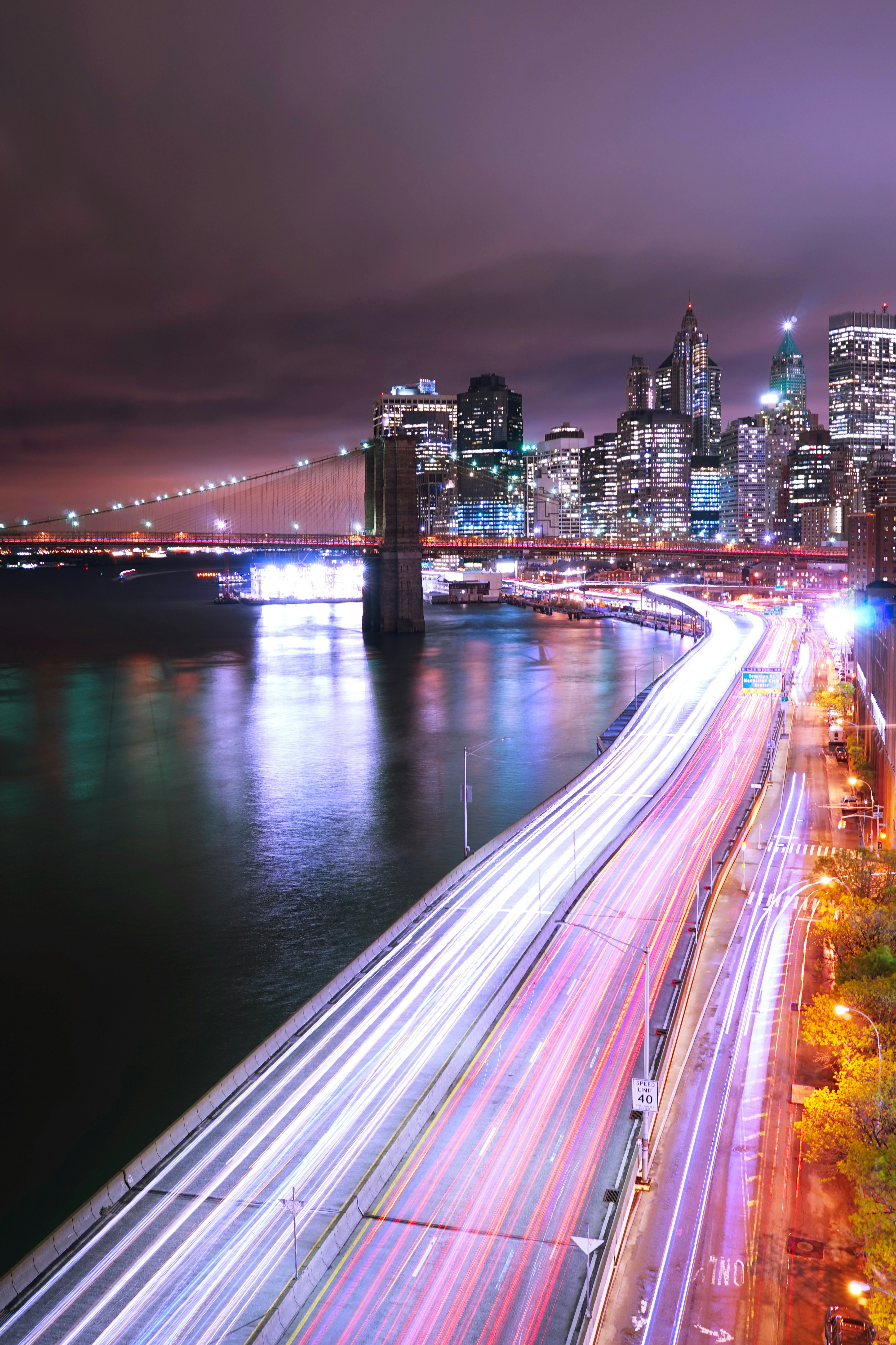 Vibrant light trails from vehicles weave through a cityscape at night, highlighting the illuminated skyline and bridge. The scene captures the dynamic energy of urban life.