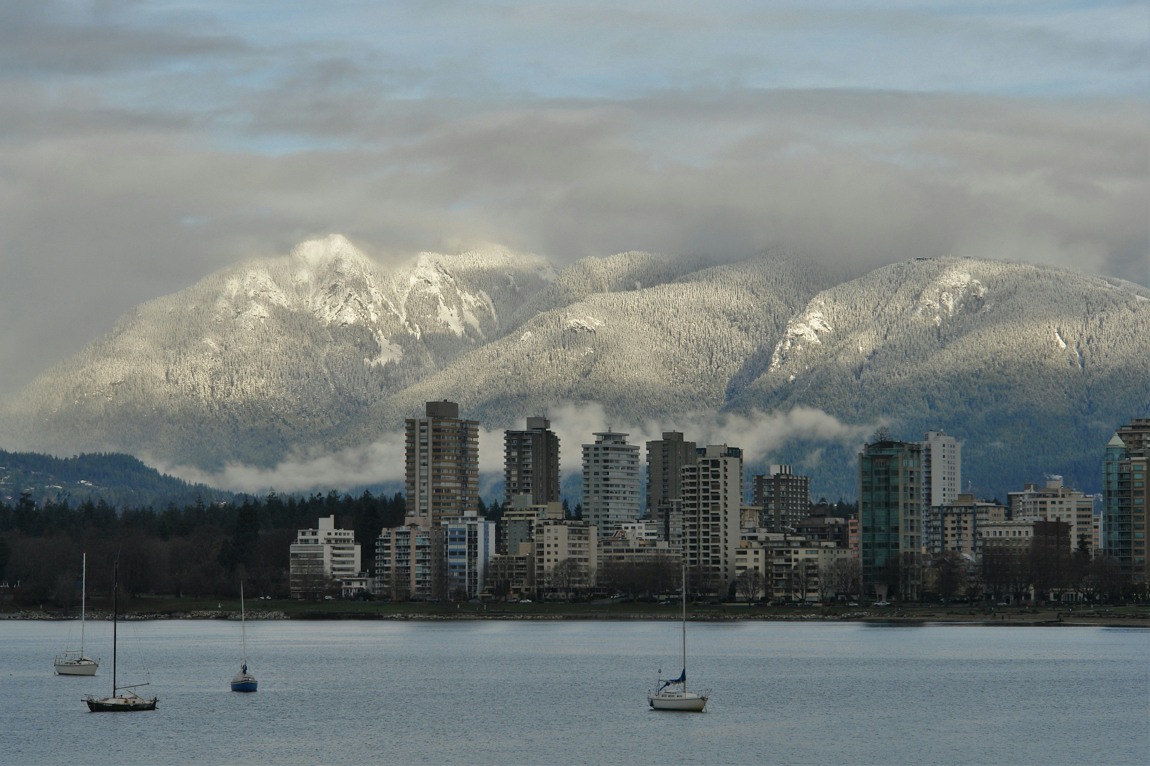 This captivating image captures a tranquil cityscape set against the majestic backdrop of snow-capped mountains. The composition is perfectly balanced with a serene body of water in the foreground, dotted with sailboats, leading the eye towards the towering city skyline. The soft, diffused lighting enhances the contrast between the urban structures and the natural beauty of the mountains, creating a harmonious blend of colors and textures that is visually striking and peaceful.