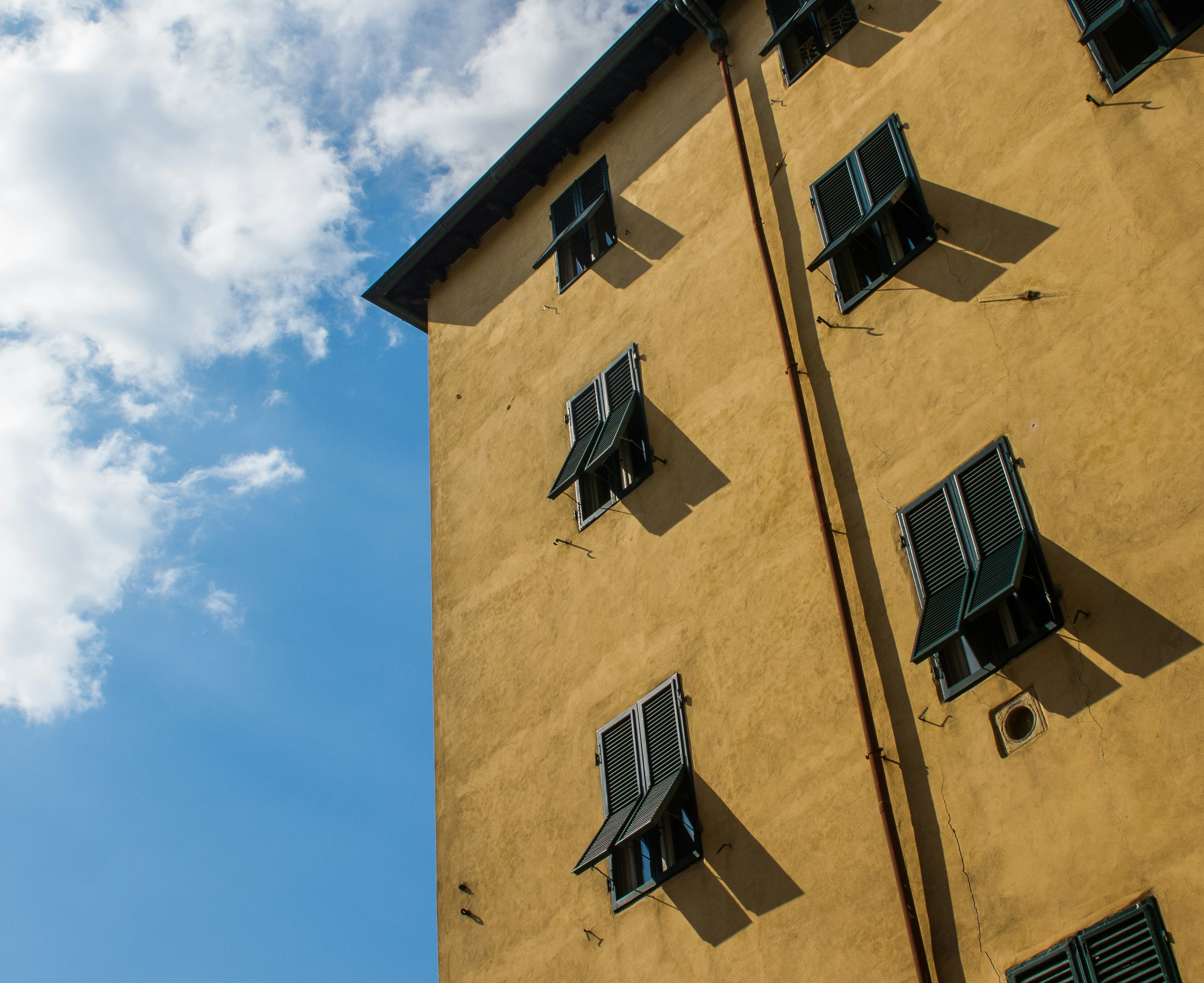 A tall building with green shutters casting shadows against a vibrant yellow facade under a bright blue sky.