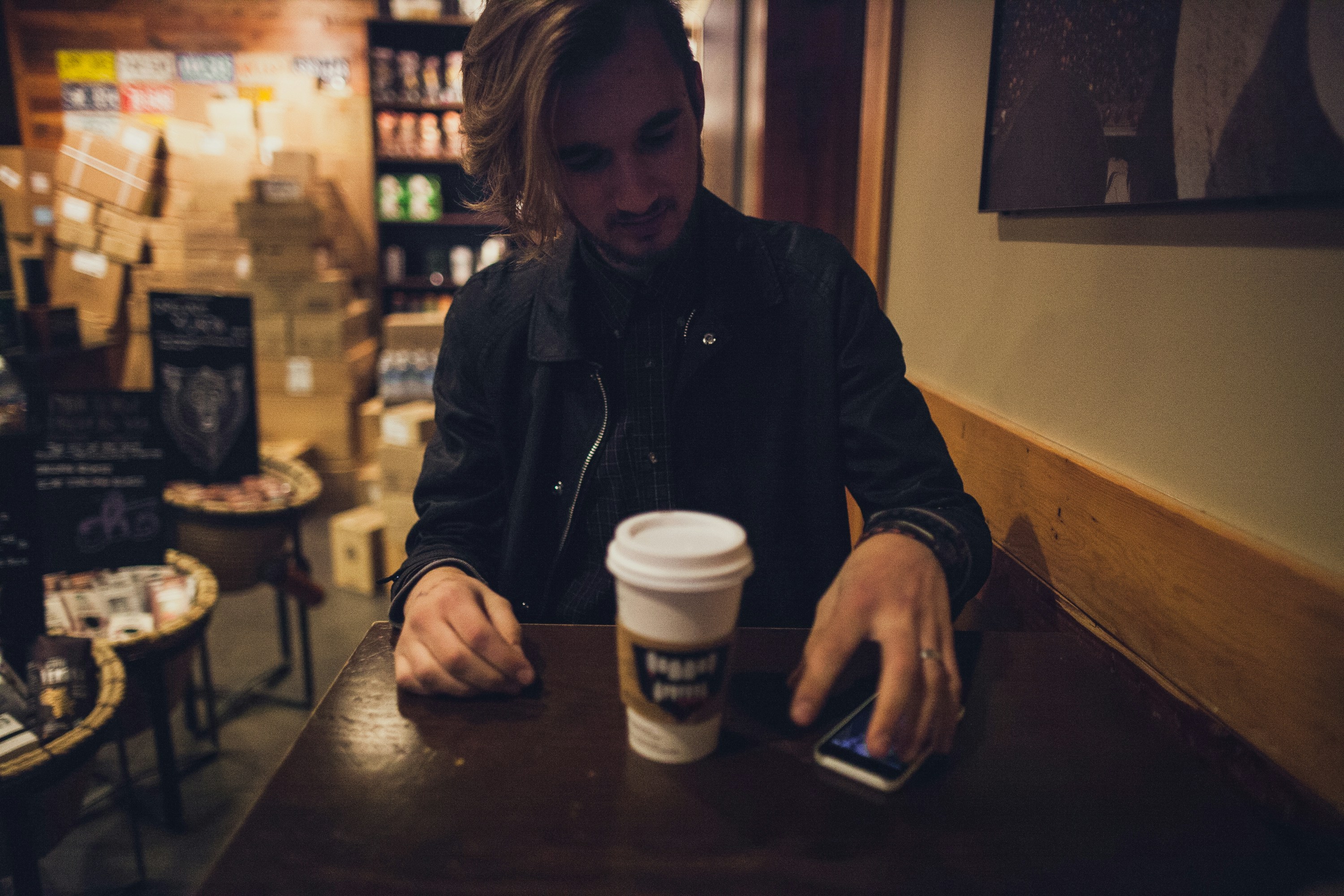 Man seated at a wooden table in a cozy café, interacting with his smartphone while a coffee cup sits in front of him.