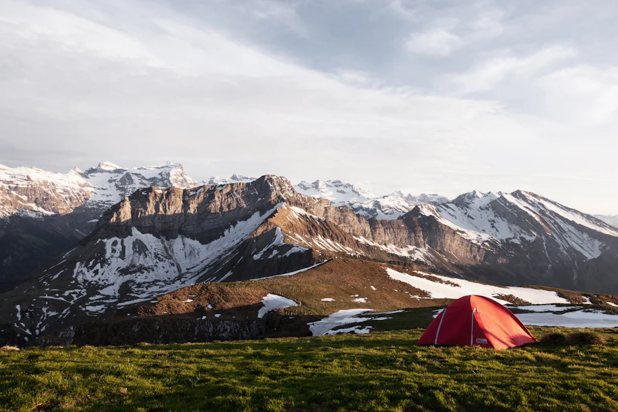 A red camping tent sits on a grassy ridge overlooking snow-streaked mountains under a pale, cloudy sky.