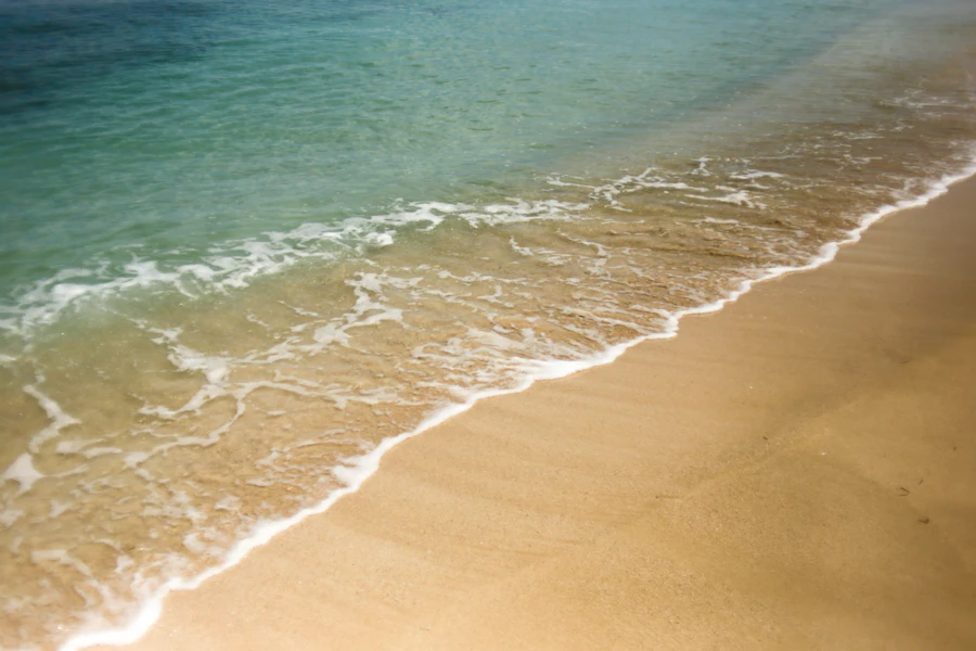 Small waves wash onto a golden sandy beach beside calm turquoise water. A diagonal line of foam traces the shoreline.