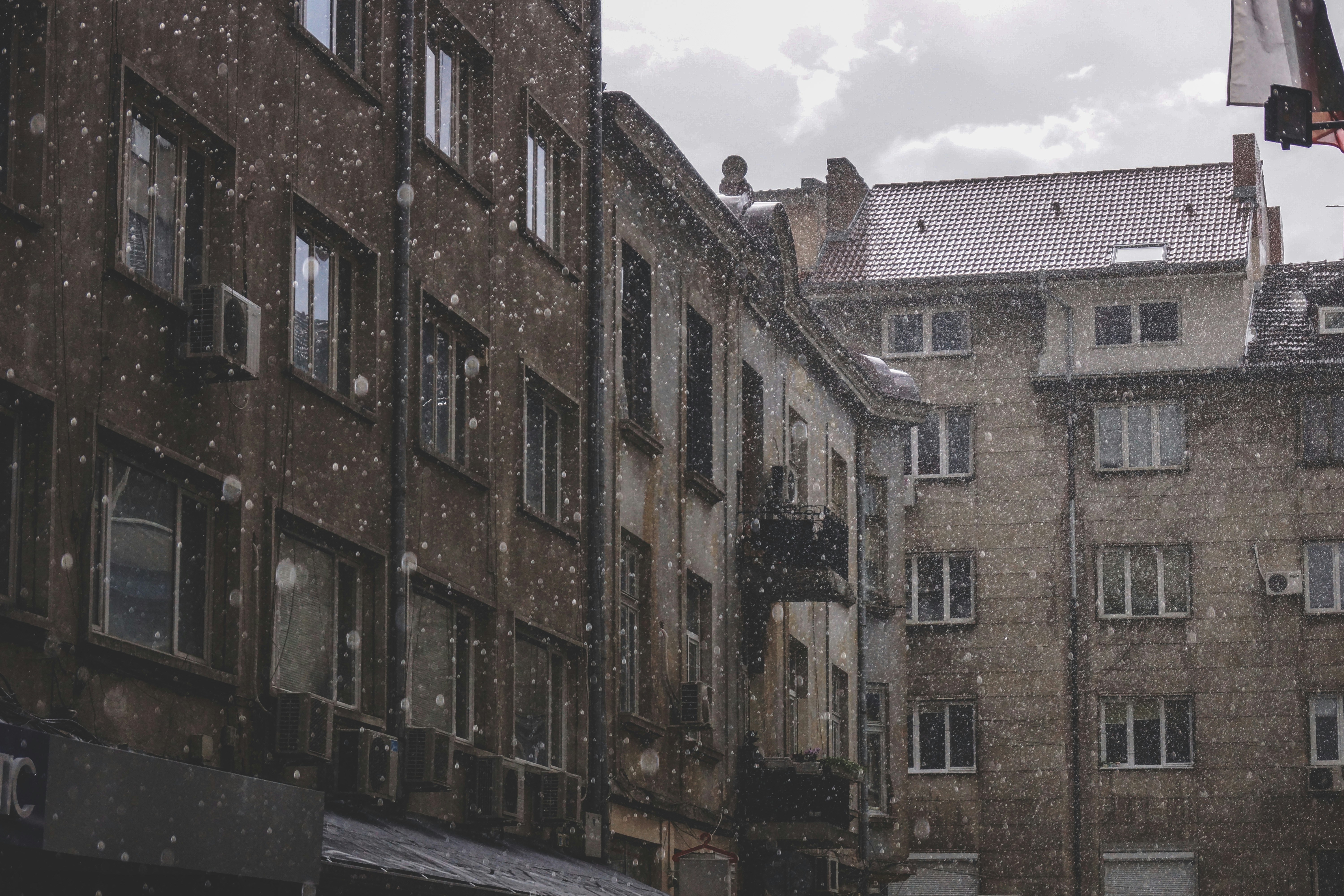 Snowflakes cascade gently between weathered urban buildings under a cloudy sky.