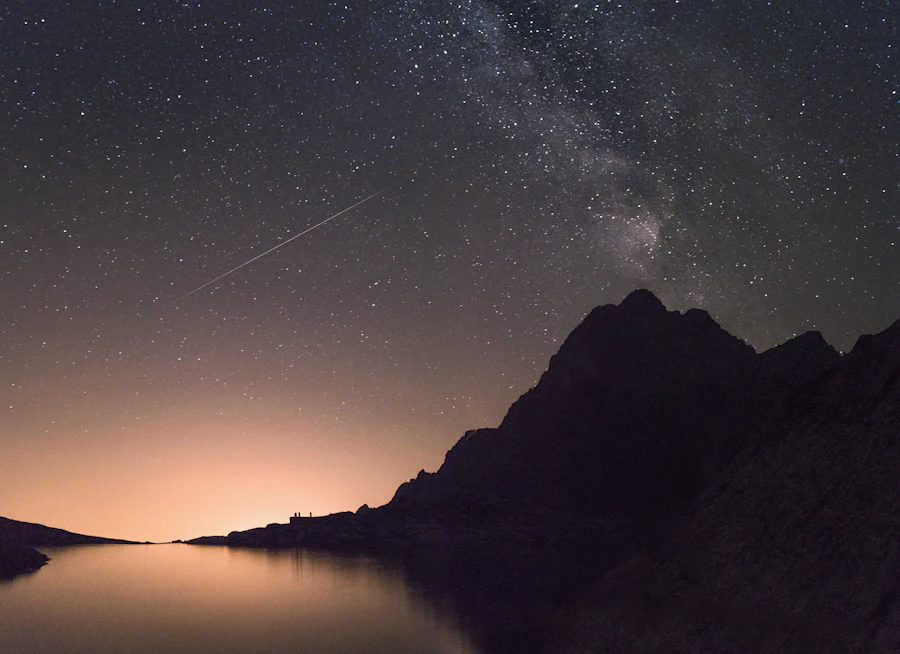 Star-filled night sky with the Milky Way arcing above dark mountain silhouettes and a calm reflective inlet below. A warm glow lights the horizon while a faint streak crosses the sky.