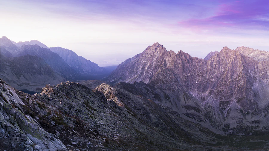 Jagged mountain peaks rise above a rocky foreground under a pastel purple-pink sky. A broad valley fades into morning haze between the ridgelines.