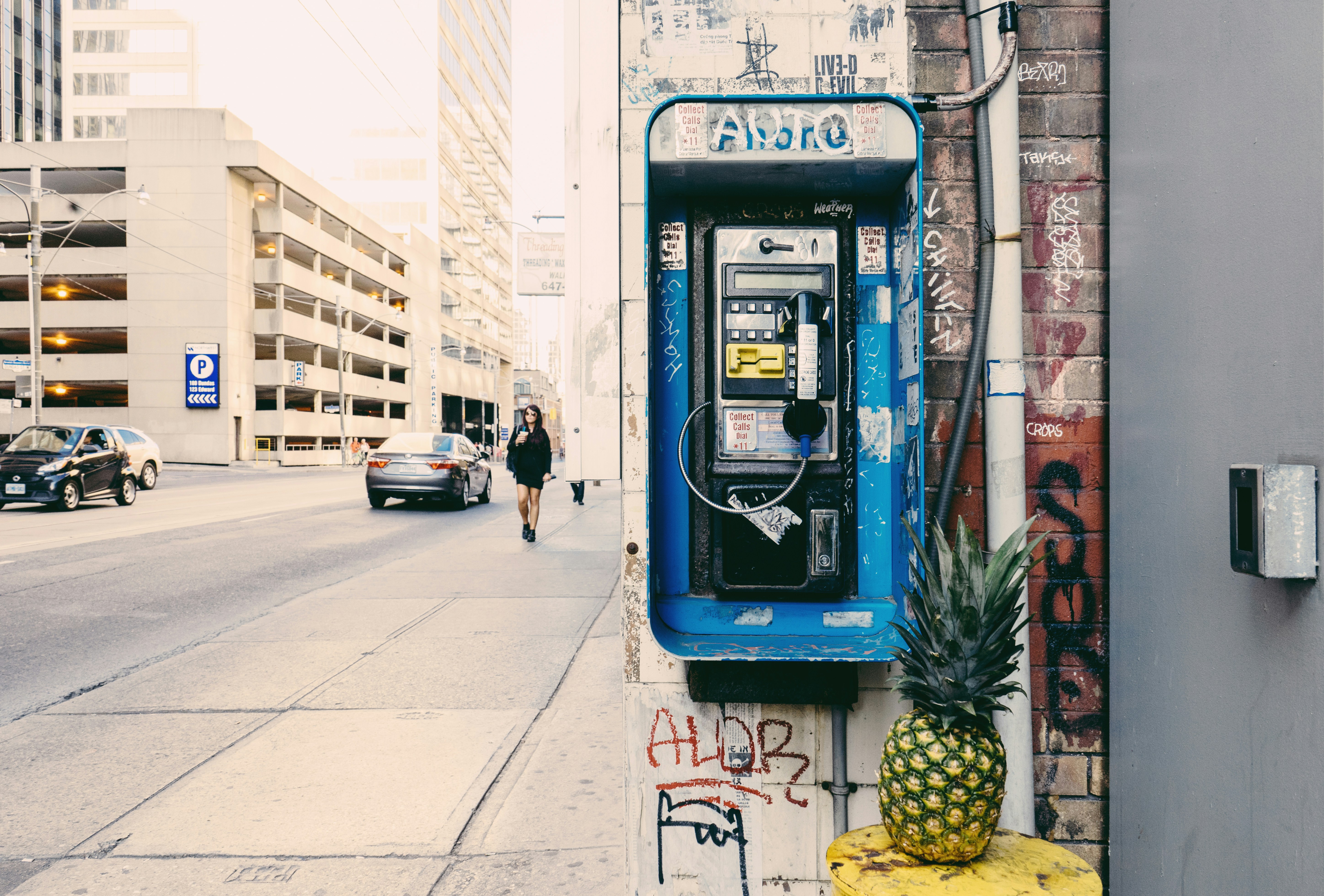 A vibrant blue payphone stands against a graffiti-covered wall, with a pineapple placed on a yellow stand nearby, capturing an intriguing blend of urban life and whimsy.