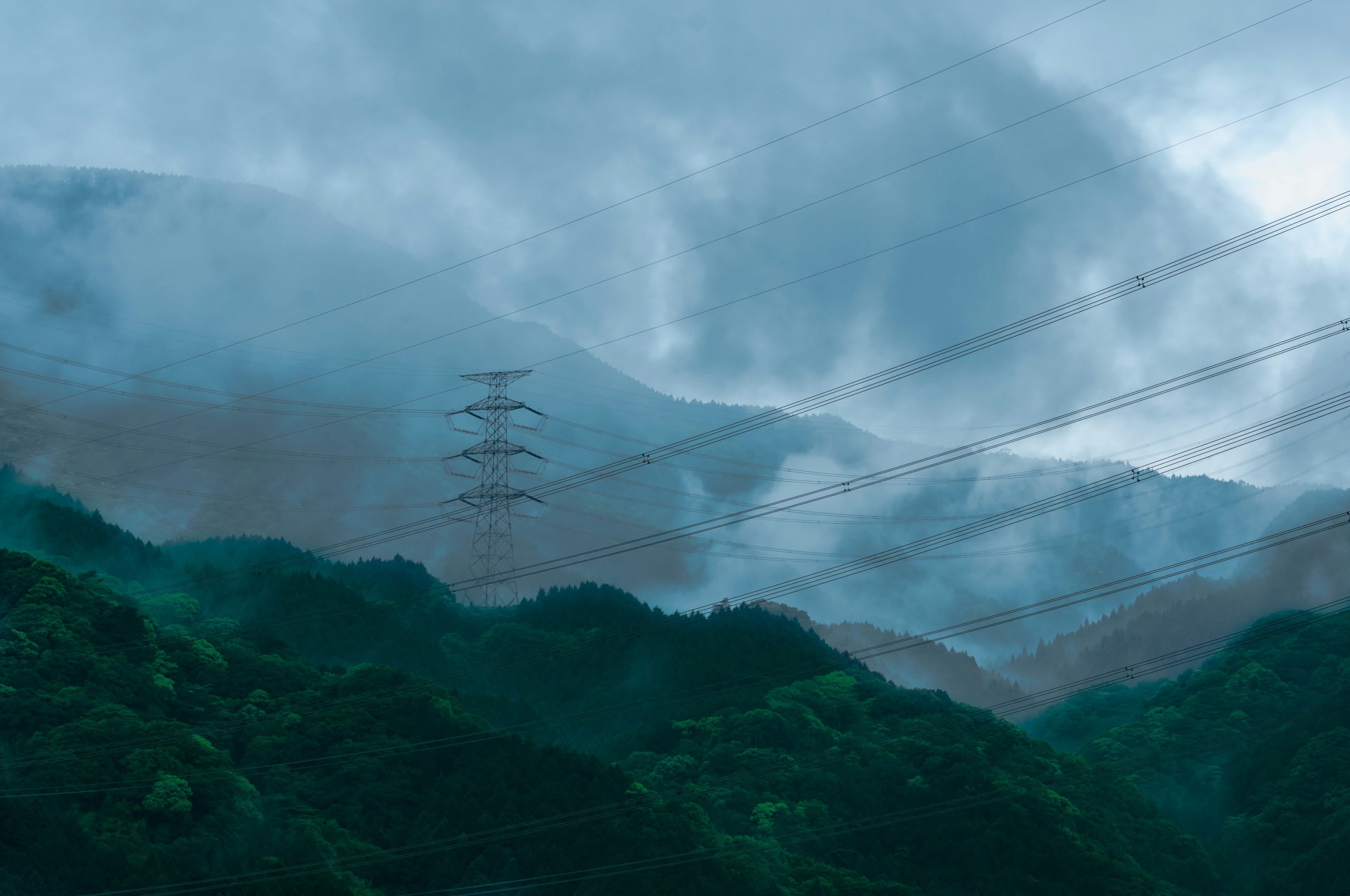 Misty mountains shrouded in clouds with electric power lines cutting through the landscape. A serene yet industrial scene unfolds.