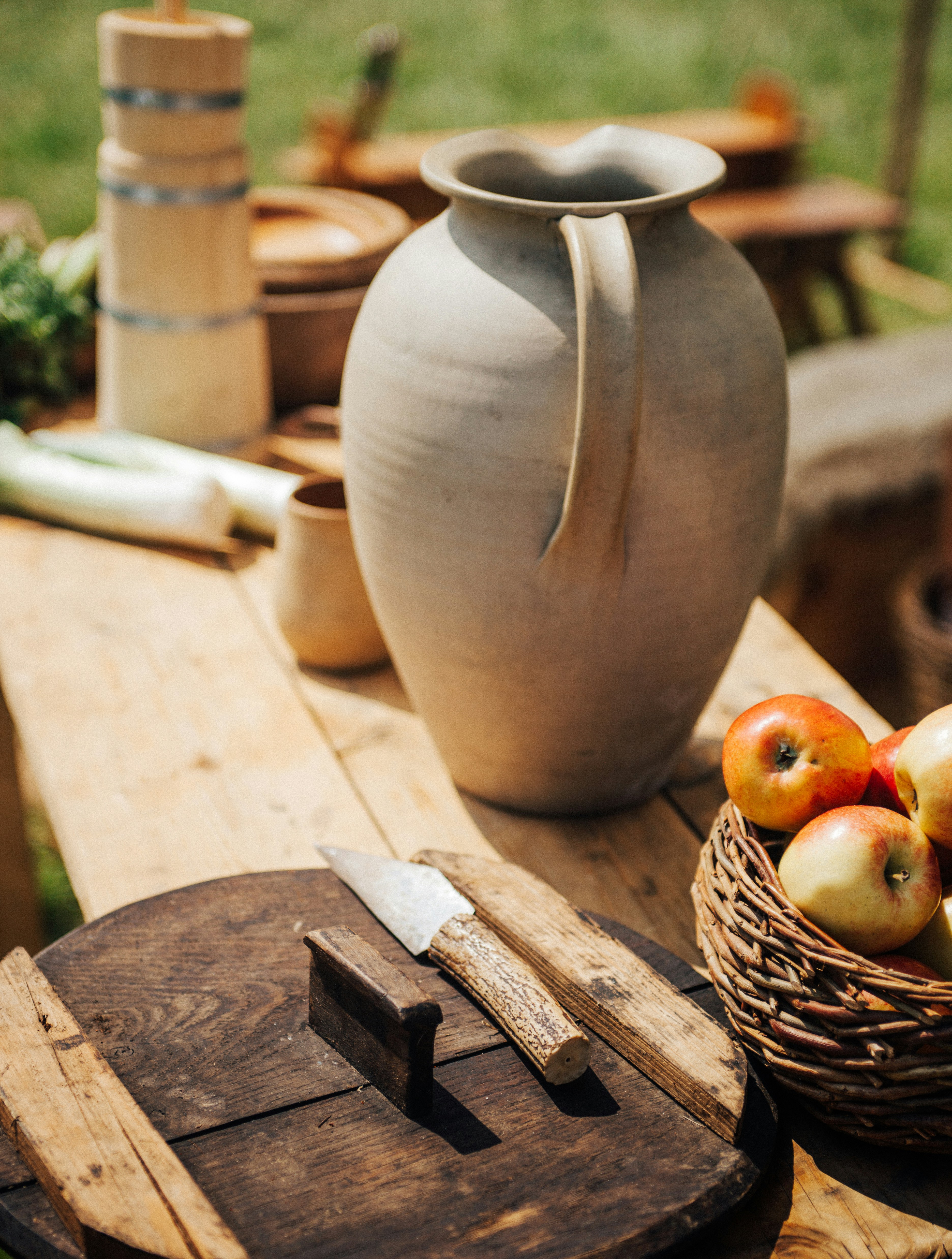 A clay pitcher and a basket of apples rest on a wooden table, surrounded by rustic kitchenware, evoking a sense of simplicity and natural abundance.