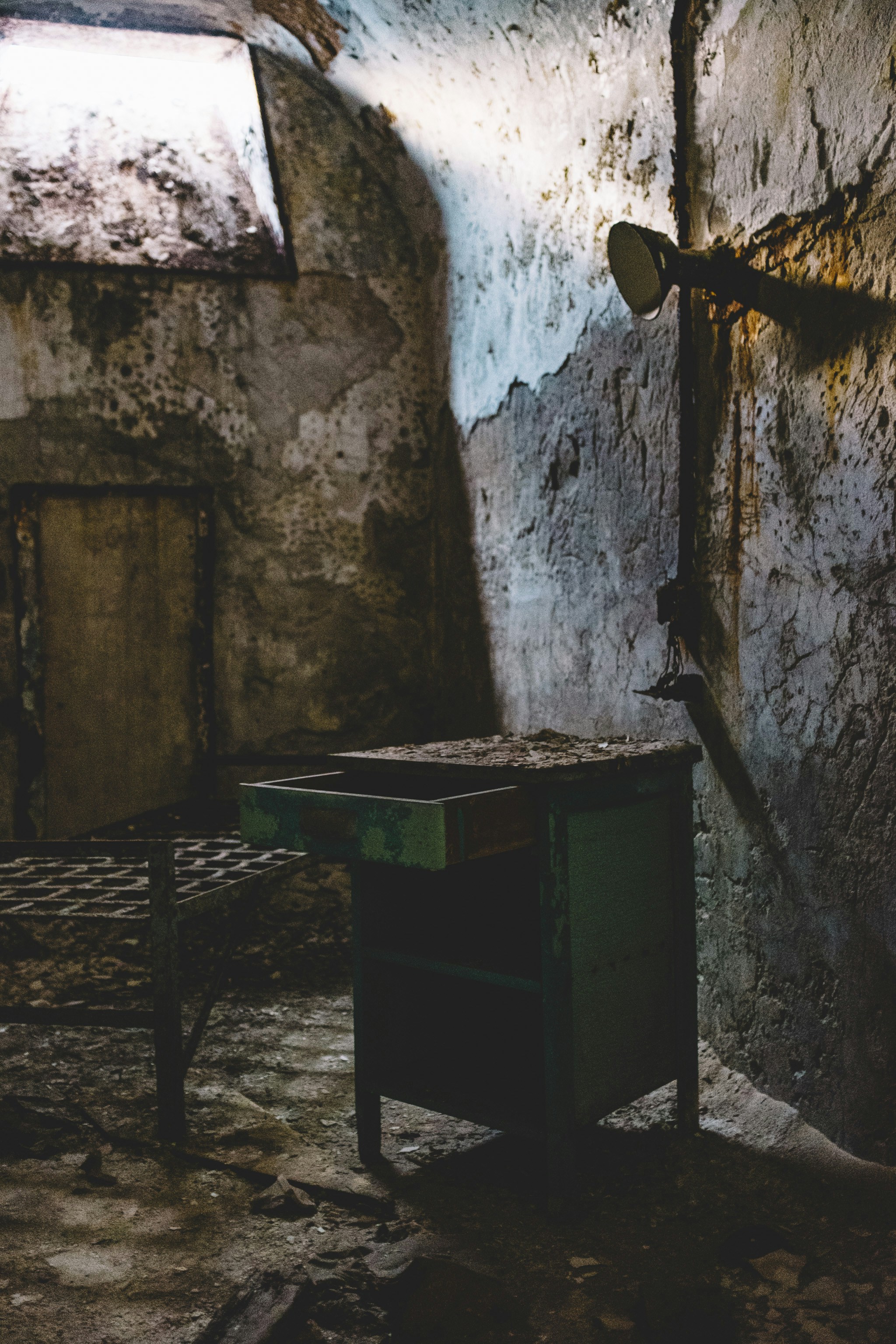 Rustic green table with a drawer in a dimly lit, dilapidated room featuring peeling walls and a faint light source. 