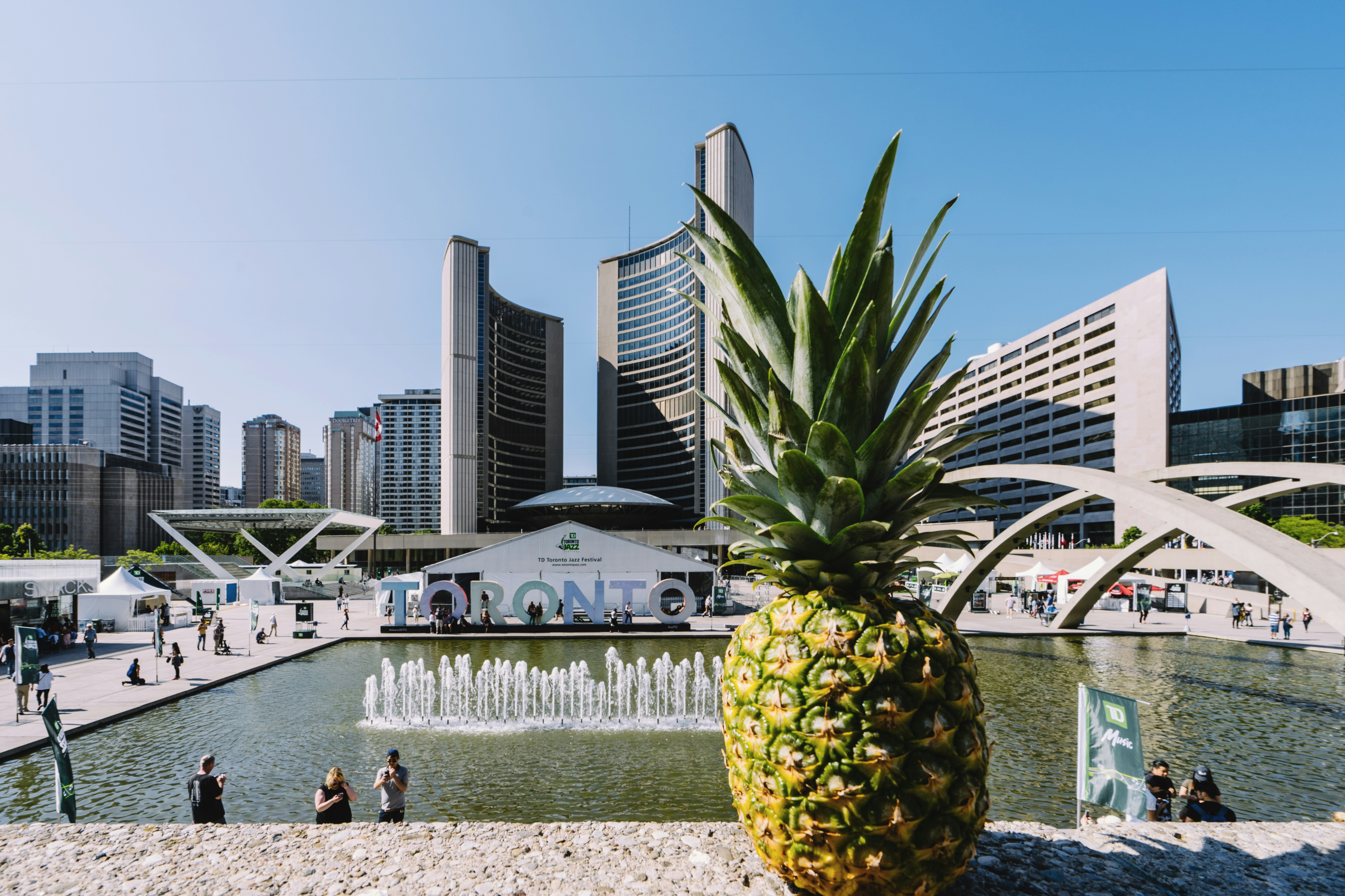 A vibrant pineapple sits in the foreground with a backdrop of Toronto's modern skyline and a bustling public square featuring a fountain.