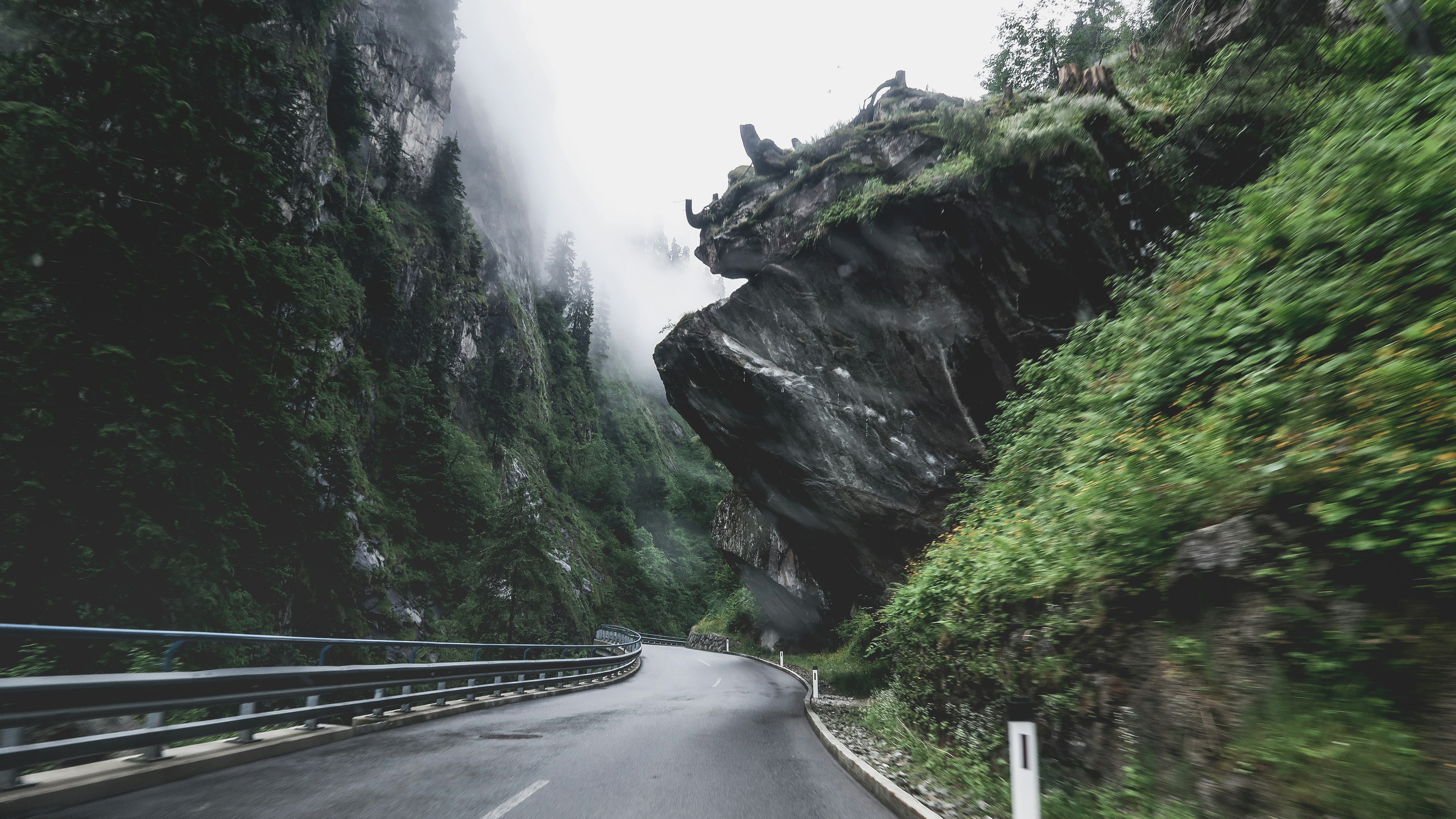 Winding road through misty mountains with lush green cliffs and overhanging boulders.