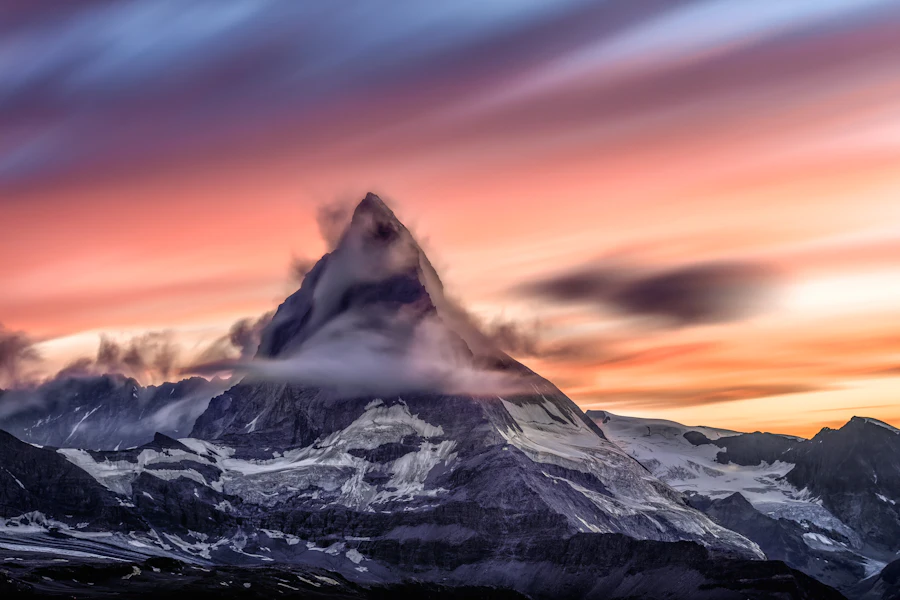 A dramatic snow-covered mountain peak rises beneath streaked clouds and a warm pink-orange sunset sky.