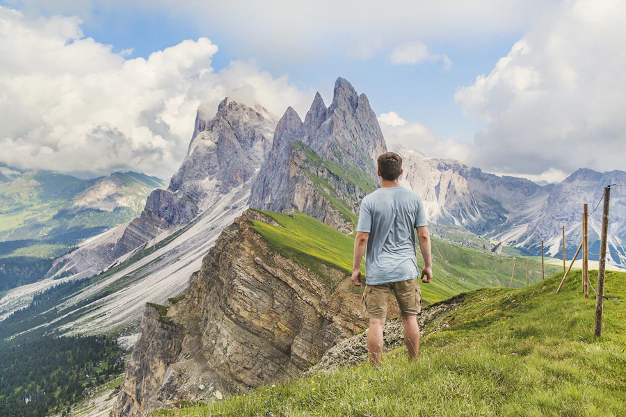 A lone hiker stands on a grassy ridge overlooking dramatic jagged mountain peaks and sweeping alpine valleys under a bright cloudy sky.