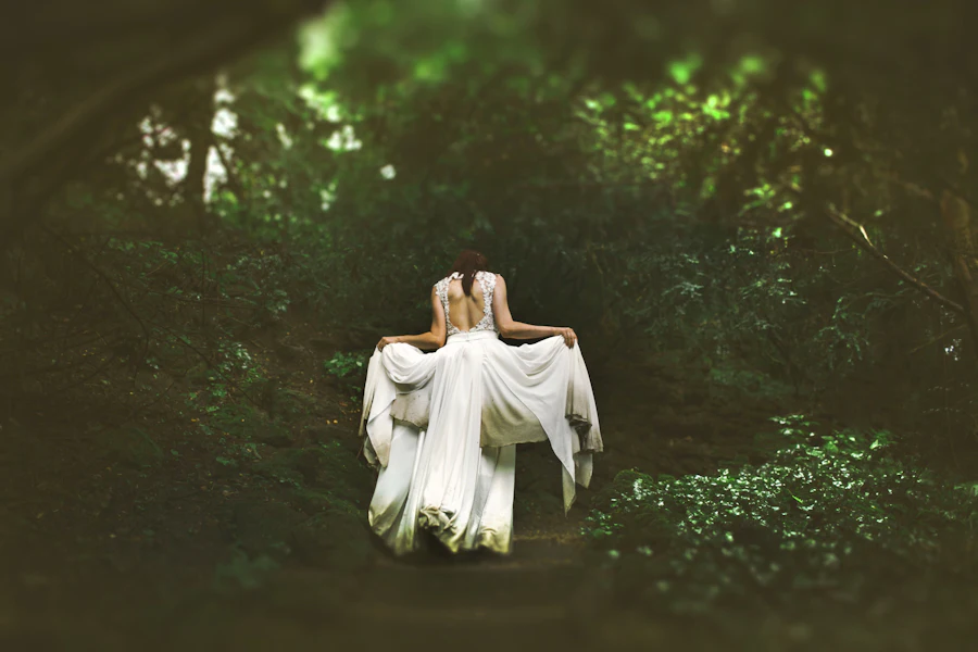 Woman in a flowing white dress walking away through a dim green forest, holding the skirt out on both sides. The scene feels soft, moody, and dreamlike.