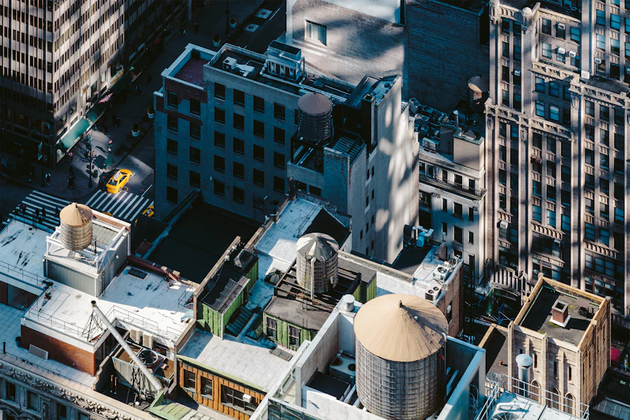 This image captures an aerial view of city rooftops, showcasing a fascinating blend of architectural forms and urban design. Dominated by cool tones and punctuated by the iconic yellow of a taxi below, the composition highlights geometric precision and the interplay of light and shadow on the buildings. The dynamic perspective and rich detail make this urban scene visually striking, evoking the bustling yet structured essence of city life.