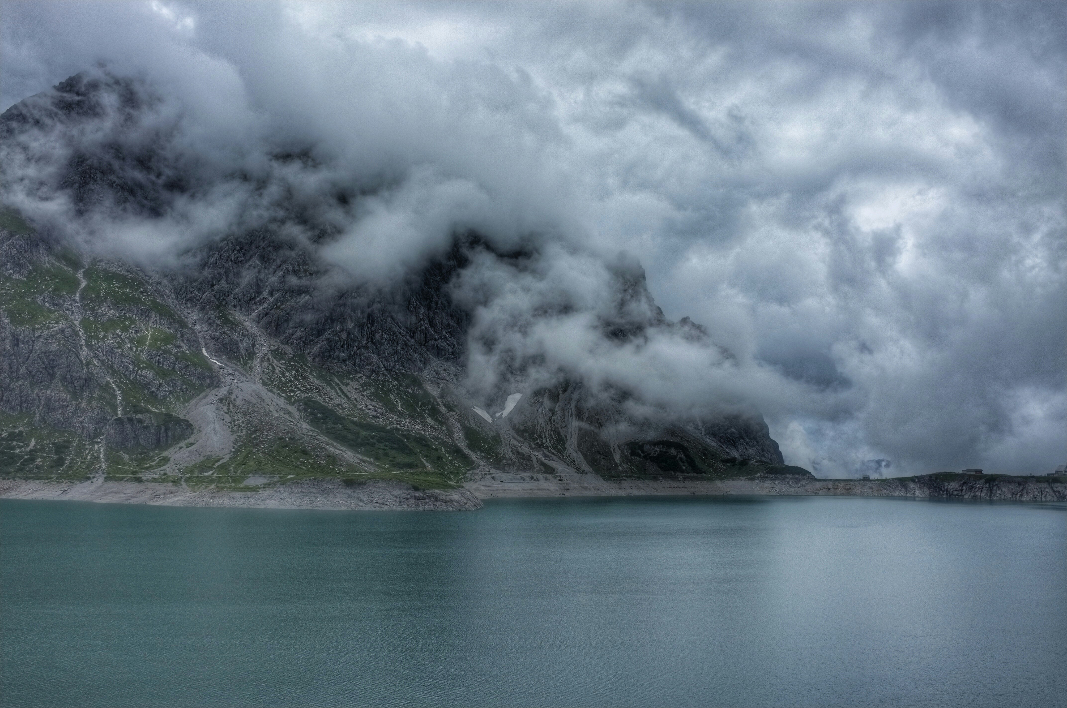 Fog envelops a rugged mountain by a calm body of water under a dramatic sky.