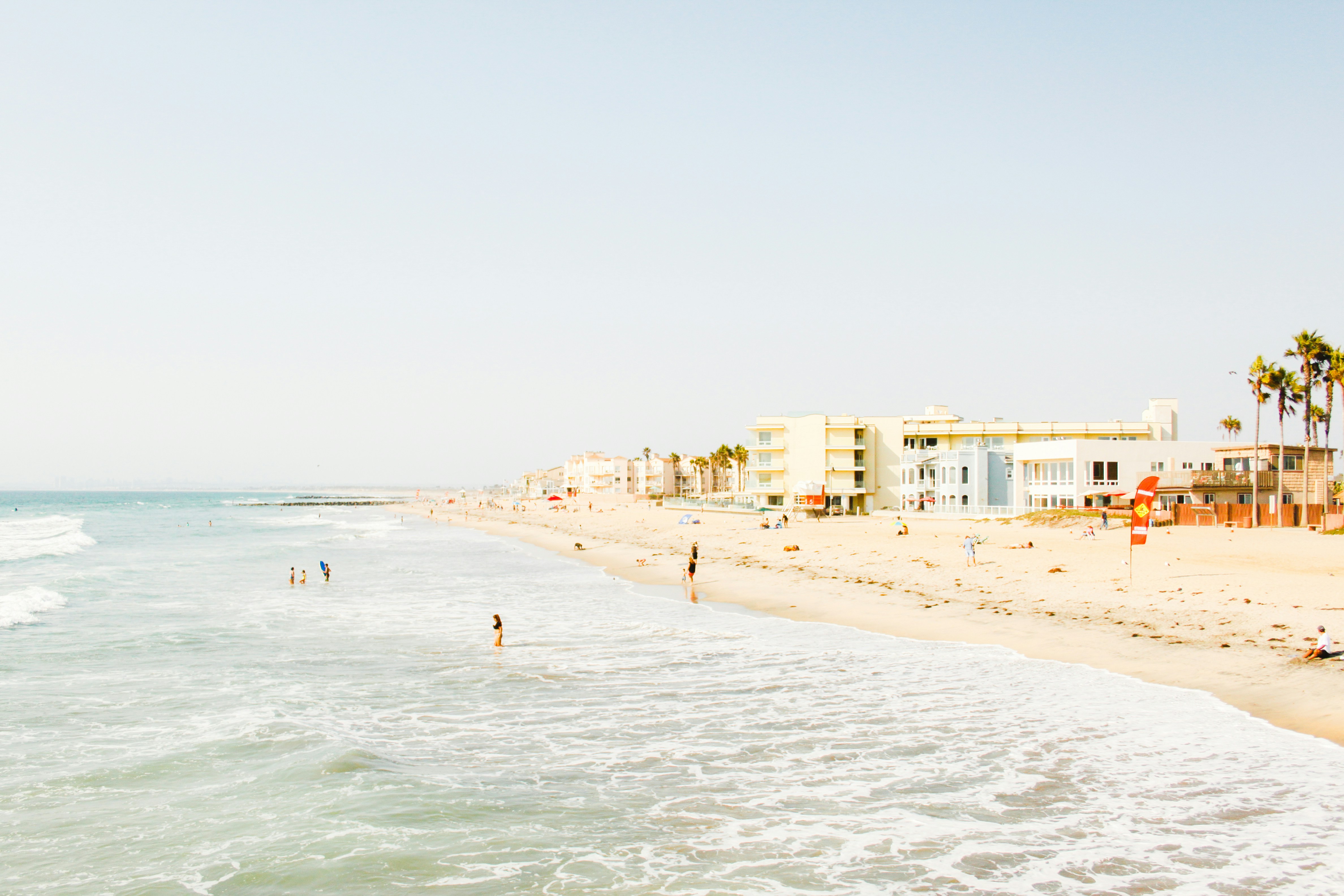 Sandy beach with gentle waves and scattered buildings under a clear sky.
