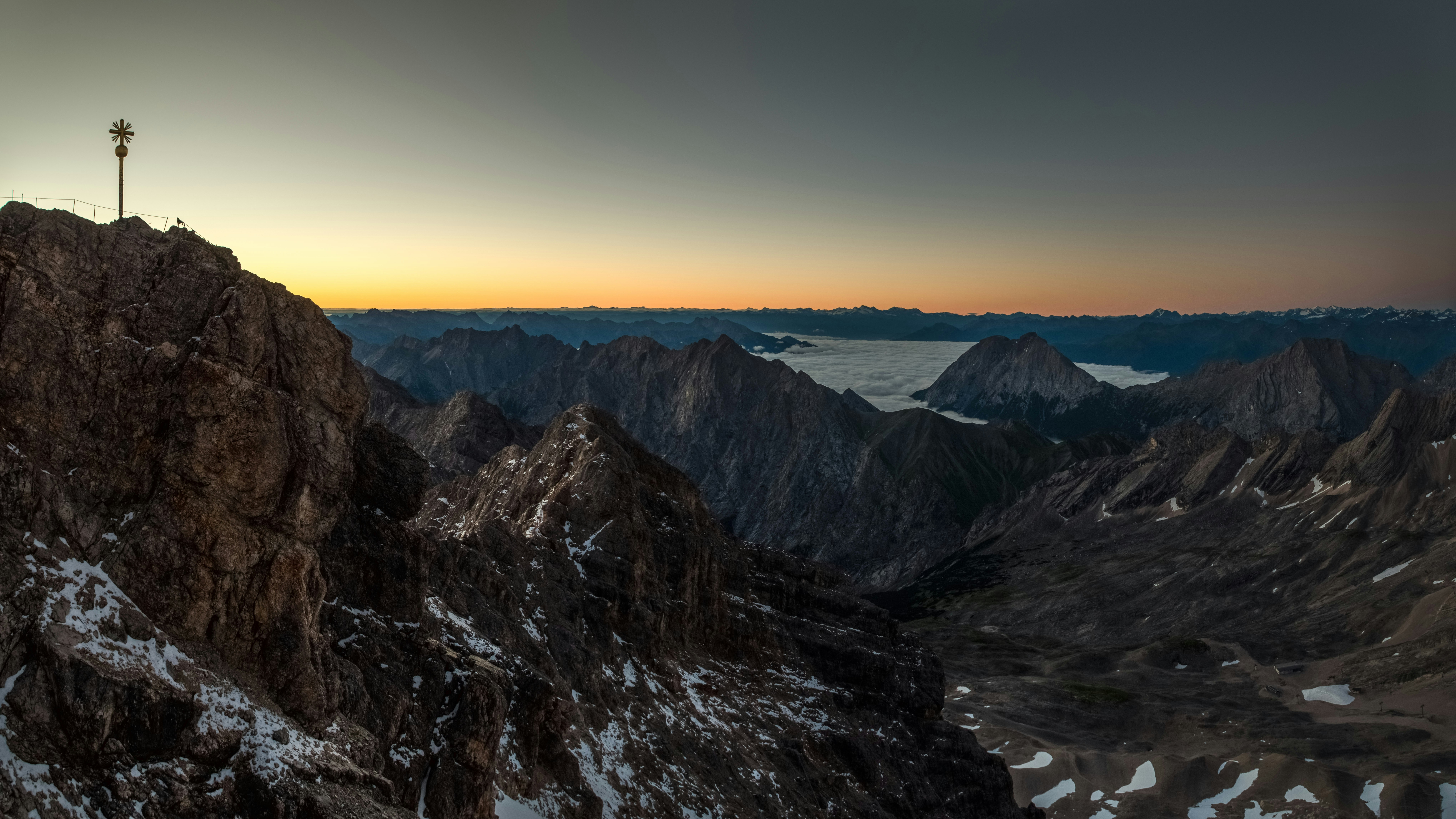 Mountain peak with cross overlooking rugged terrain and distant horizon at sunrise.