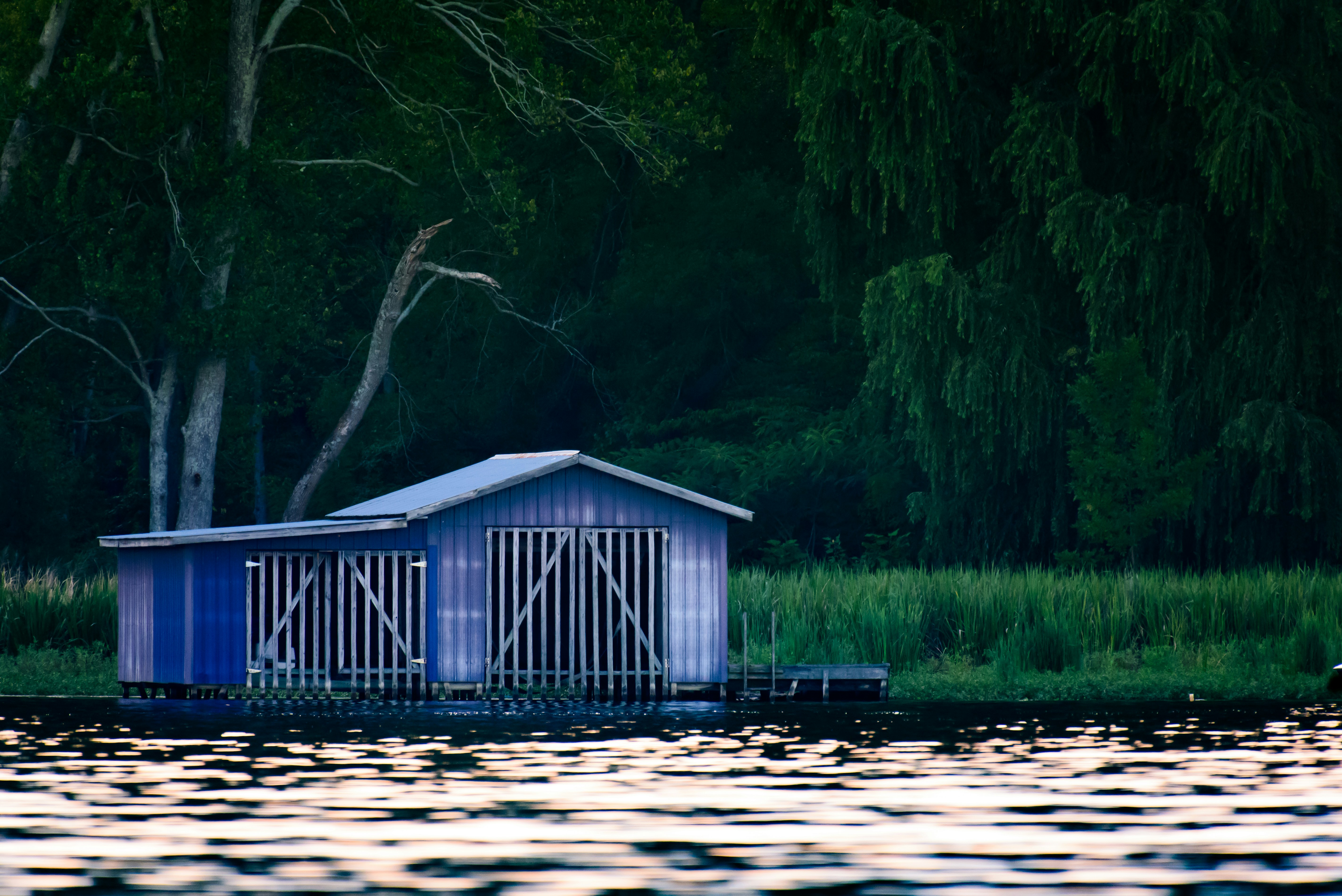 A tranquil blue boathouse stands by the serene waters, surrounded by lush greenery and softly rippling reflections at dusk.