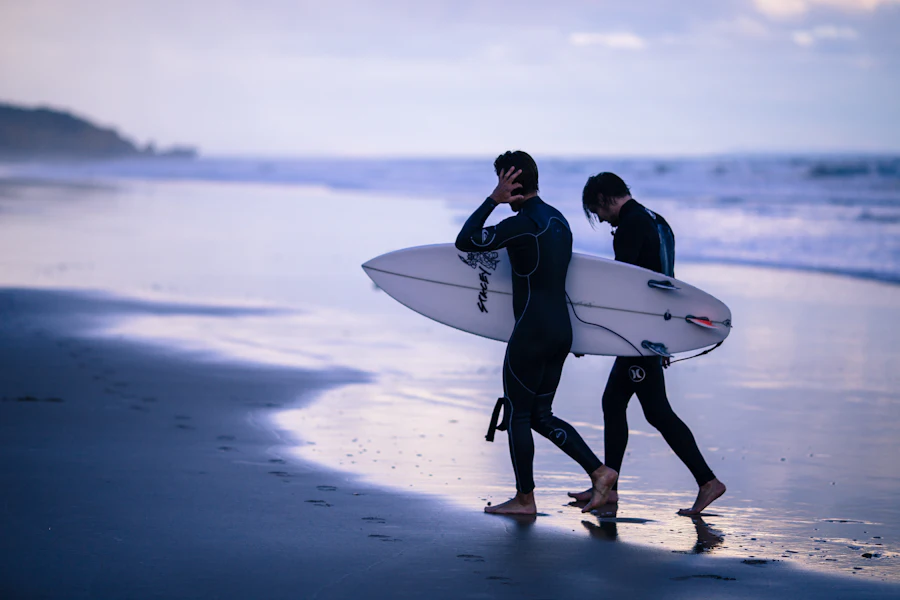 Two surfers in wetsuits walking along a sandy beach with surfboards under a twilight sky.