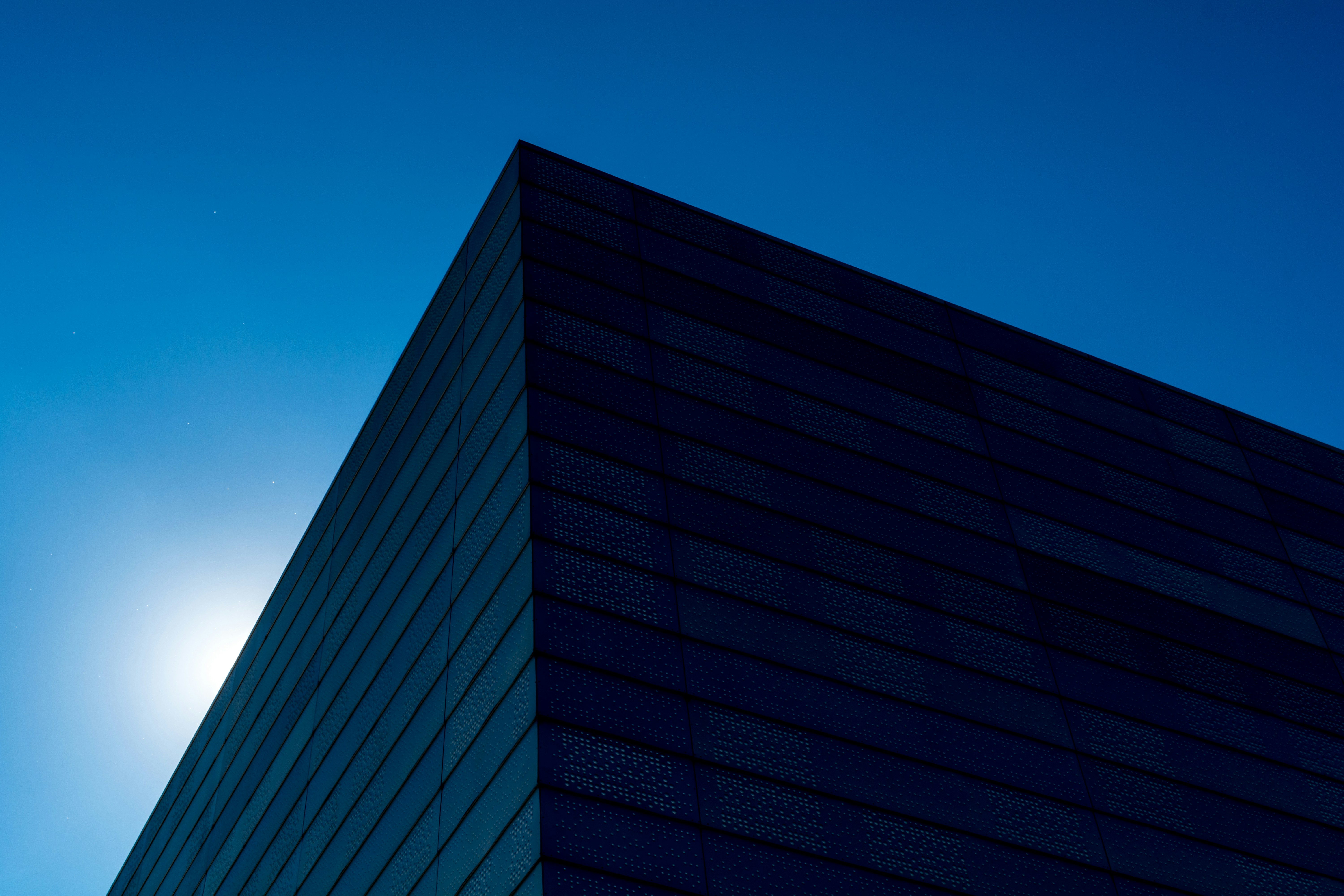 Modern building facade in silhouette against a clear blue sky with the sun peeking from behind.