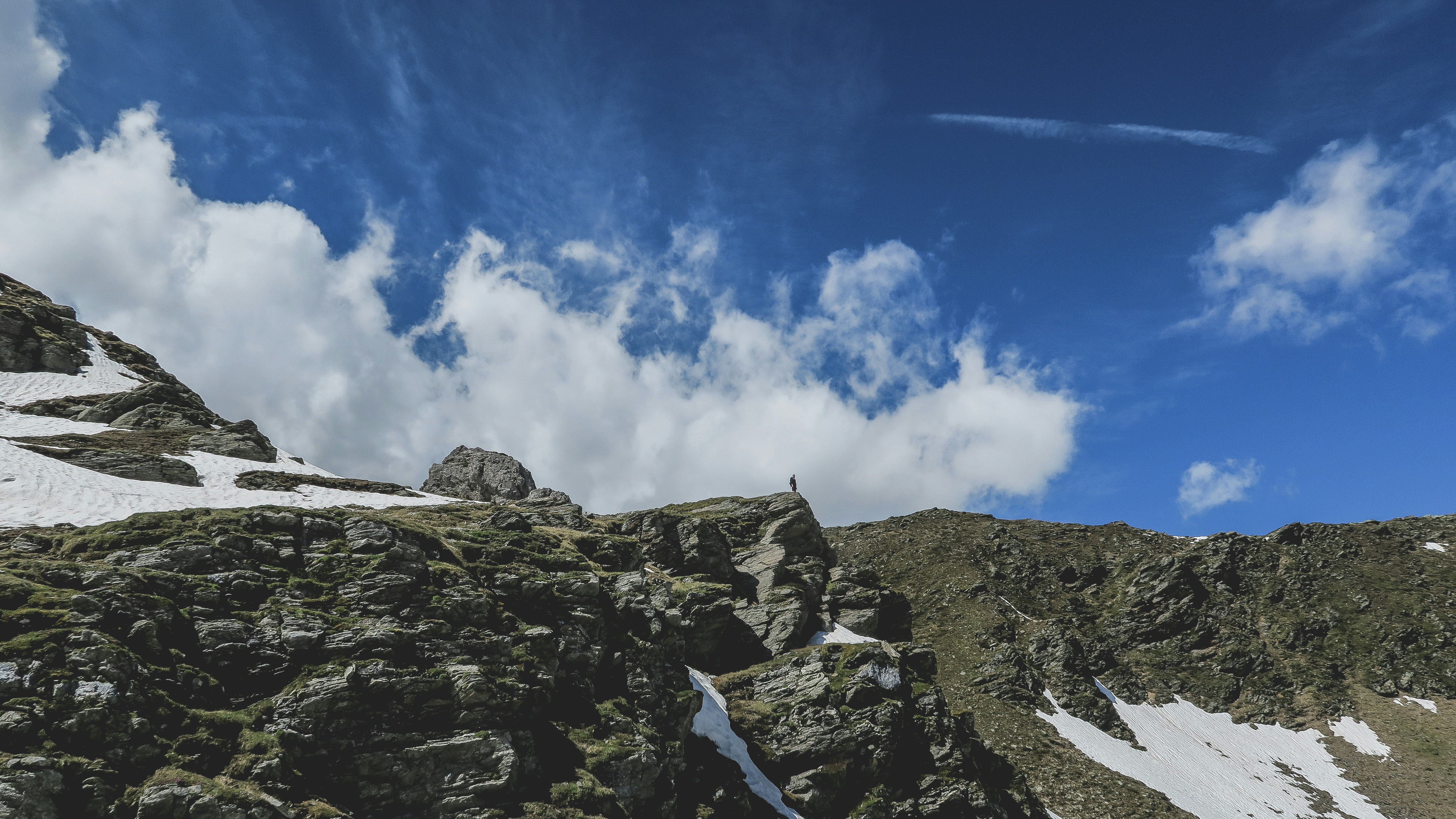 A lone figure stands atop a rocky outcrop, surrounded by rugged mountains and patches of snow under a vibrant sky.