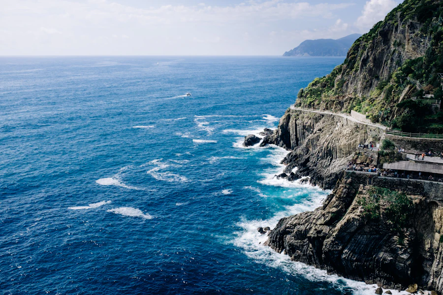A rugged coastal cliff drops into deep blue water, with white surf breaking along the rocks and a narrow road tracing the shoreline. Distant hills fade into the hazy horizon.