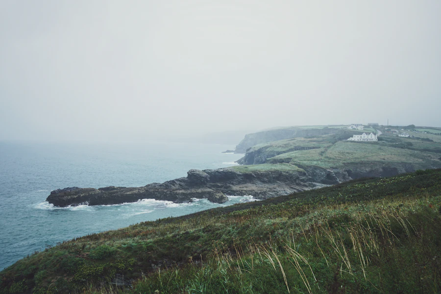 Misty coastline with dark rocky cliffs, pale blue sea, and a white house perched on the headland in the distance. Grassy foreground slopes toward the shore under a soft overcast sky.
