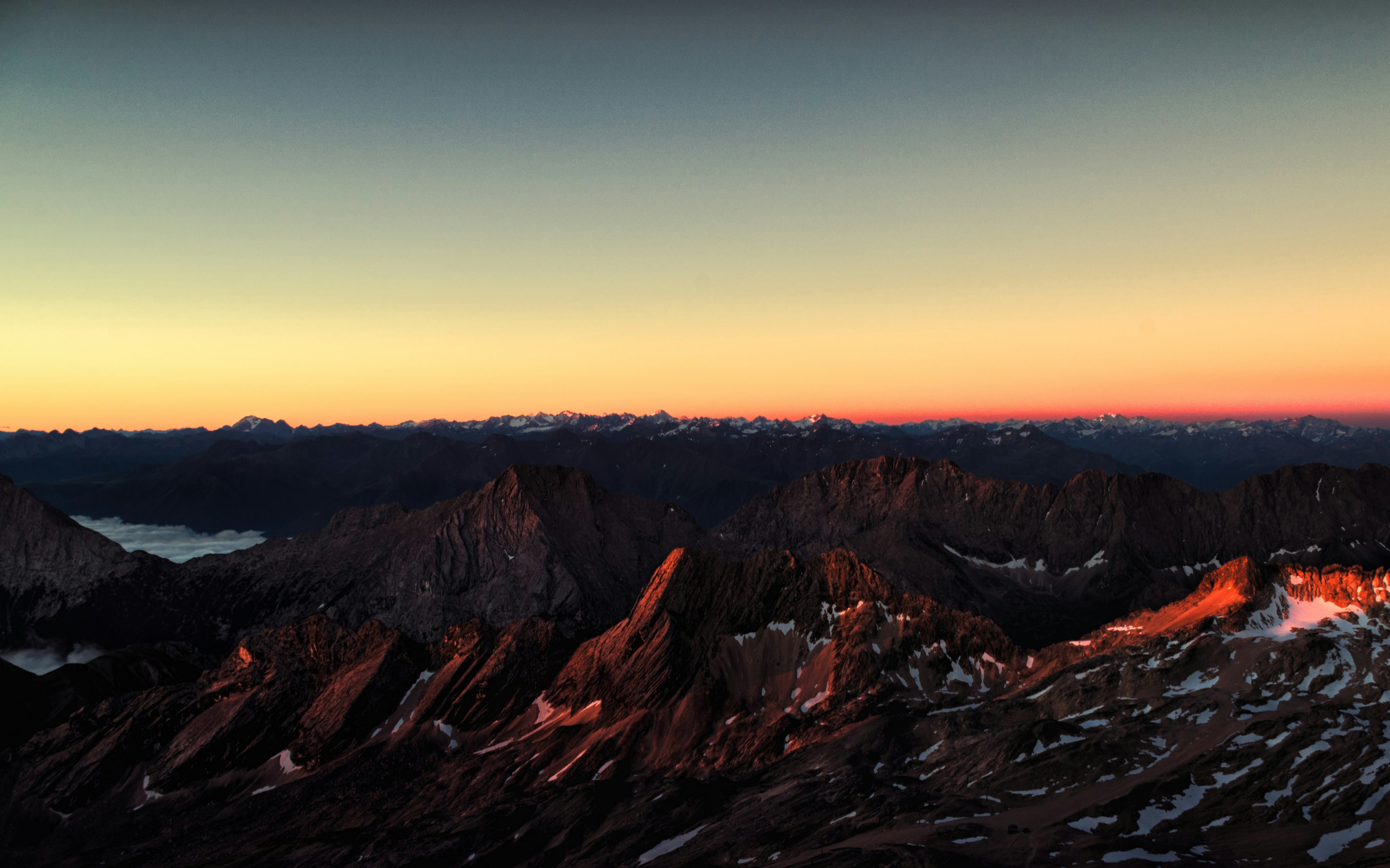 Mountain range under a vibrant gradient sky at sunset, with snow-capped peaks illuminated by warm light.
