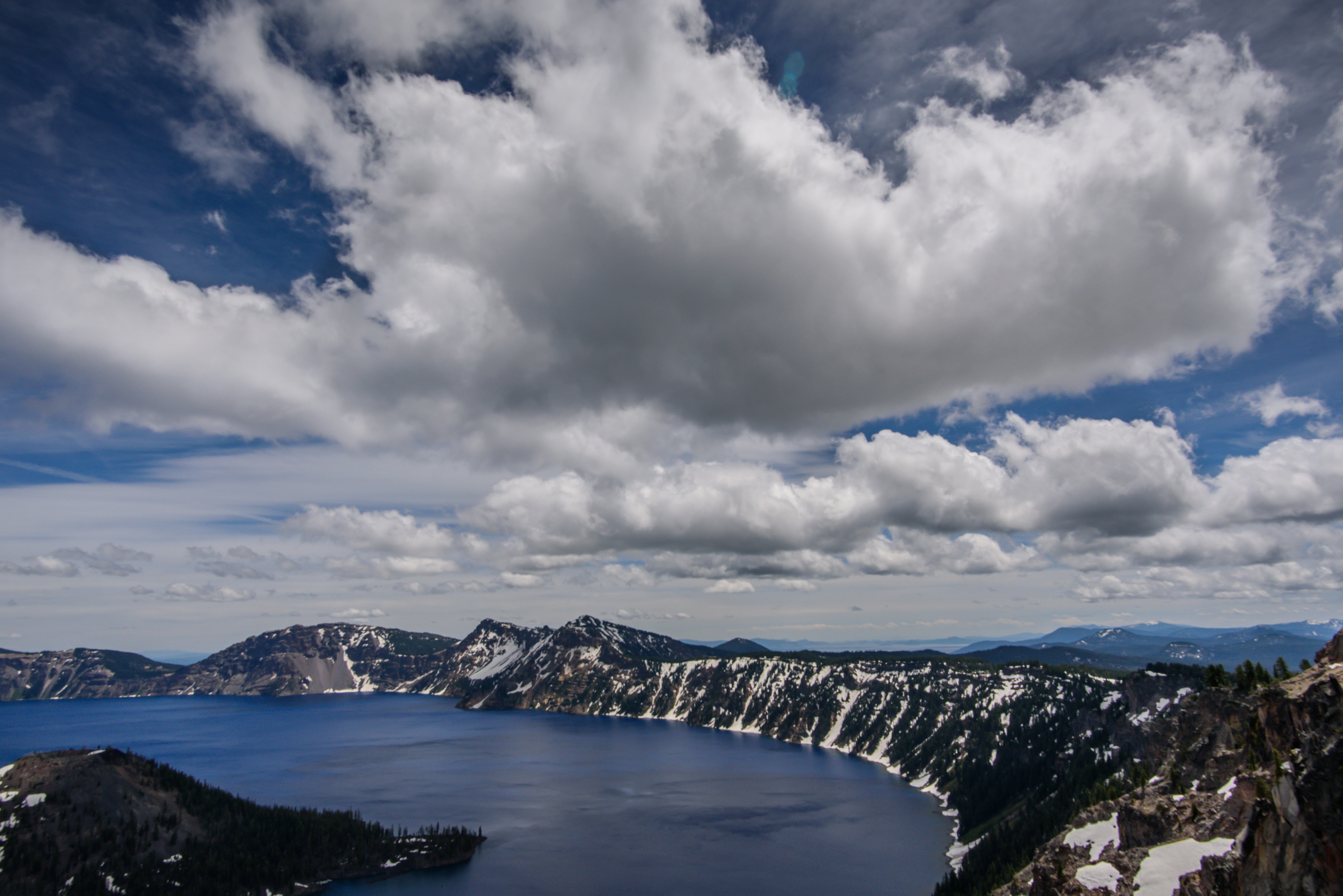 Vast blue waters of Crater Lake framed by rugged cliffs and dramatic cloud formations. Snow remnants hint at the changing seasons.