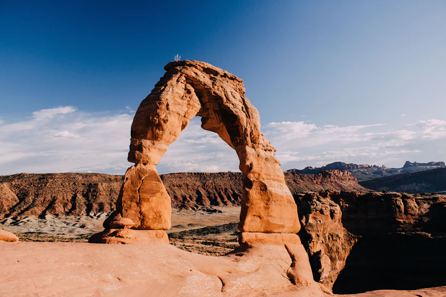 A sandstone arch stands against a blue sky above a rugged desert landscape, lit by warm late-day sun. Distant mesas and a deep shadowed drop-off frame the scene.