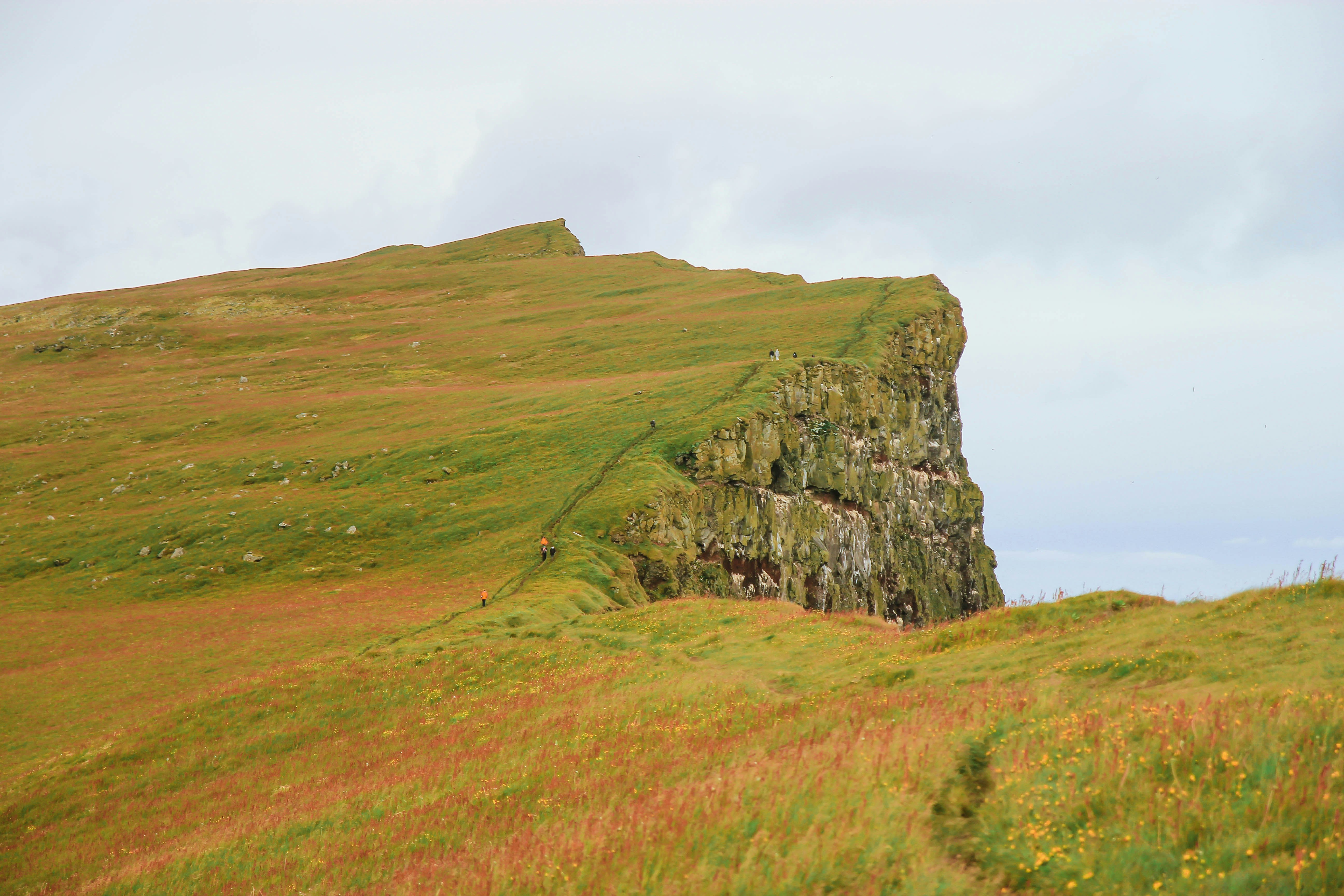 Lush green hills meet a rugged cliff edge under a cloudy sky, showcasing the natural beauty of the landscape. A few hikers can be seen along the trail, emphasizing the scale of the terrain.