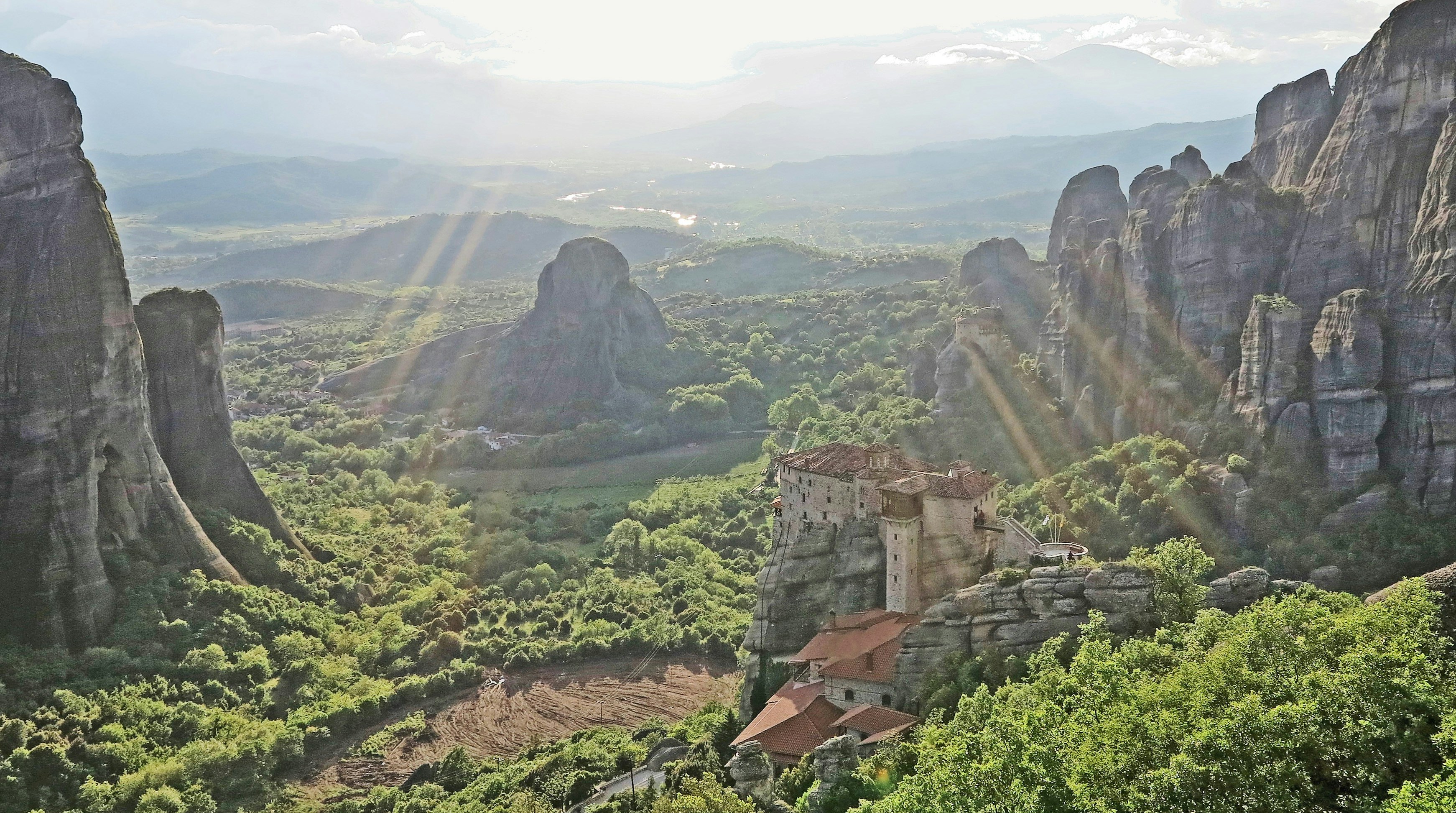 Monastery nestled among towering rock formations with sunbeams streaming through the sky in Meteora, Greece.