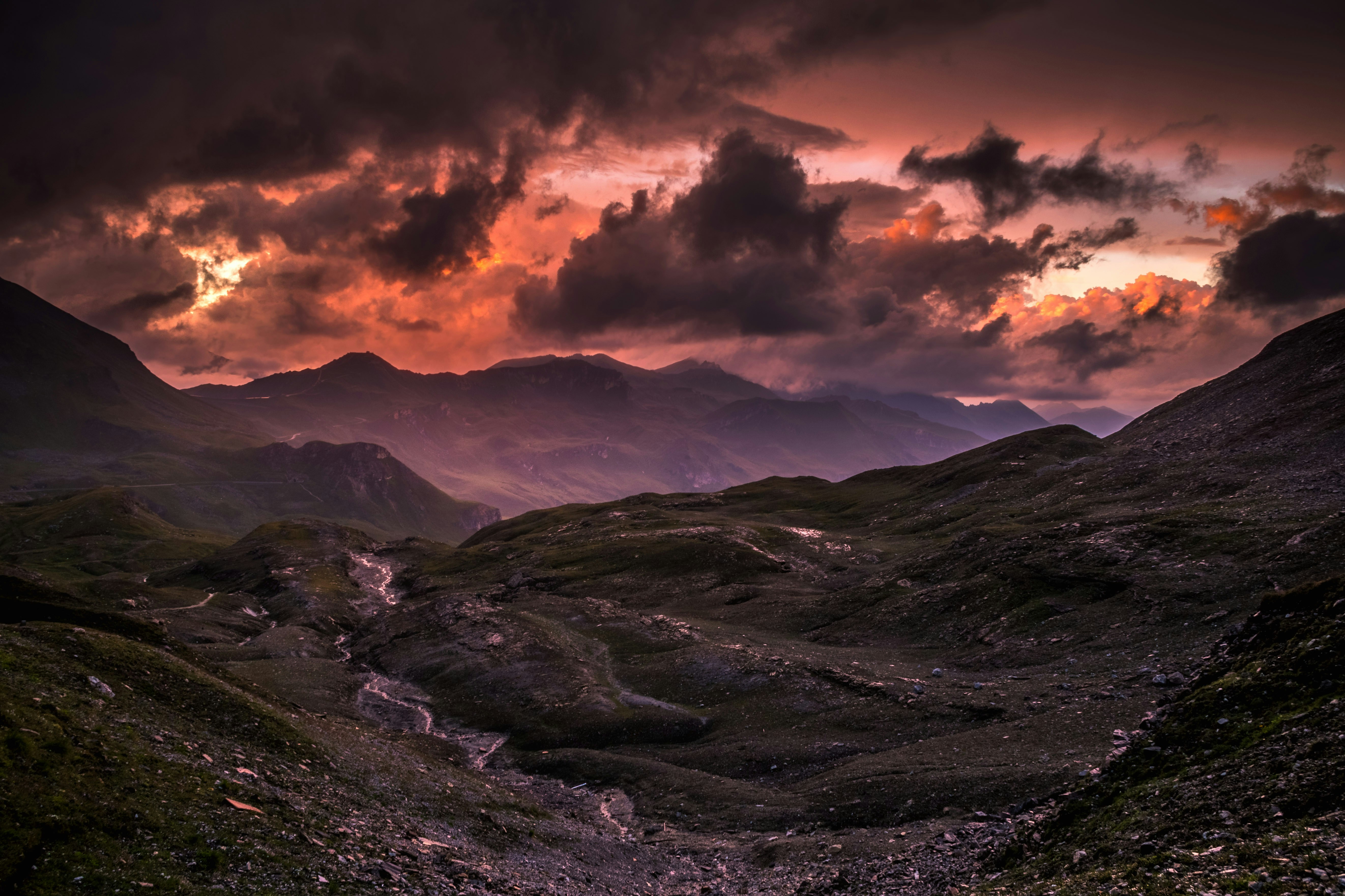 Dramatic sunset casts a fiery glow over a mountainous landscape with rolling hills and ominous clouds.