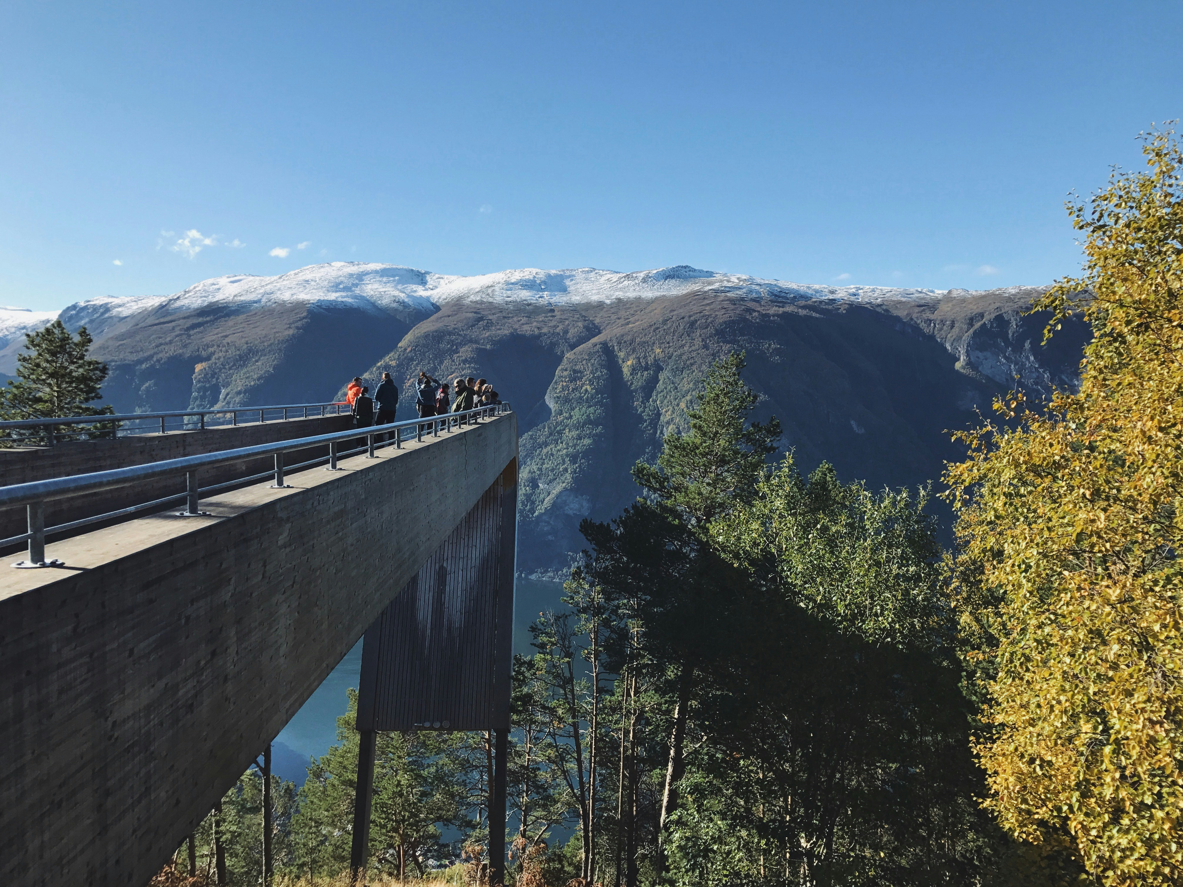 Visitors stand on a cantilevered viewpoint overlooking a serene fjord, framed by lush greenery and distant snow-capped mountains.