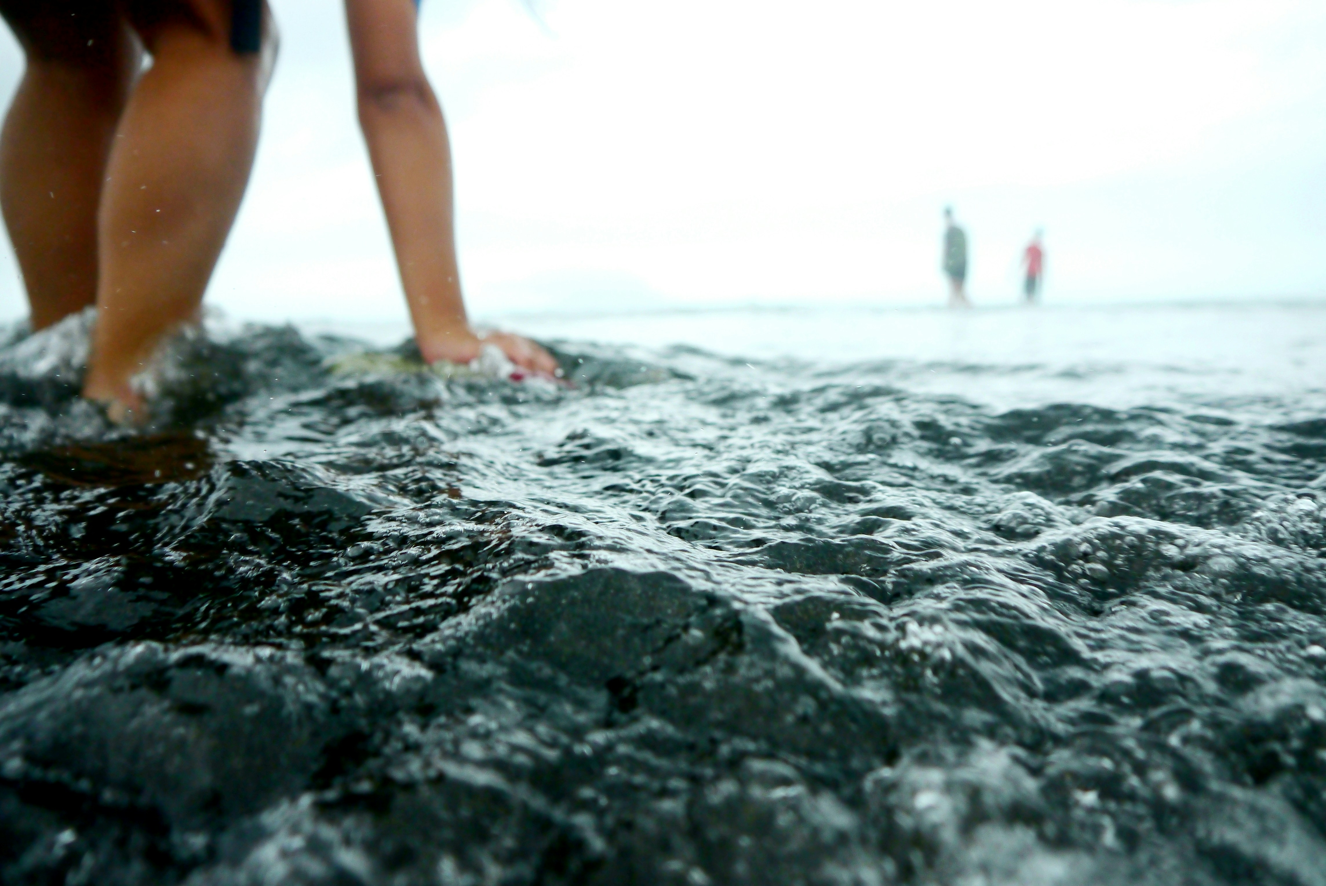Close-up of a person wading through shallow water with blurred figures in the background on a foggy beach.