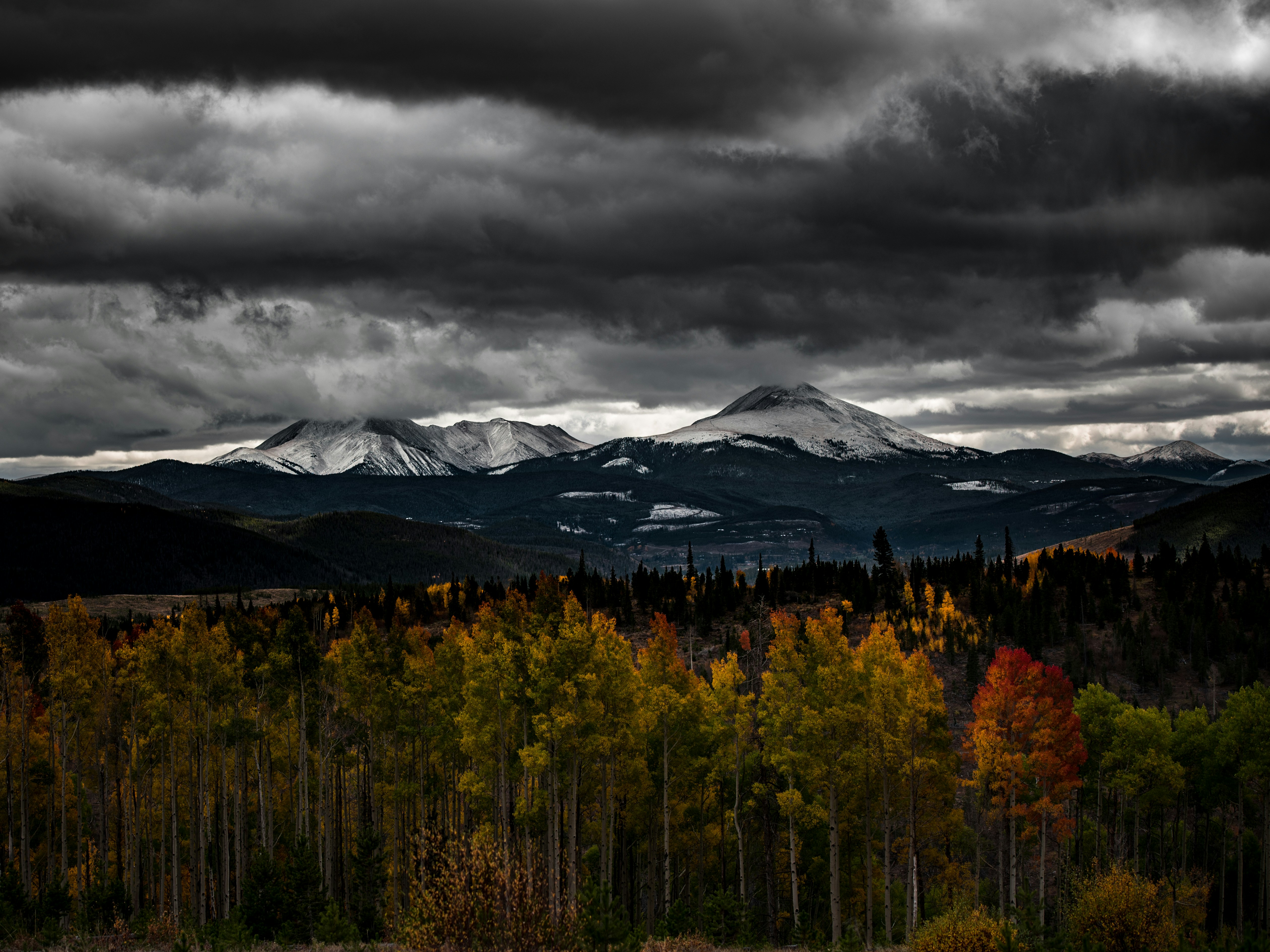 Vibrant autumn foliage in the foreground contrasts with the snow-capped mountains and dramatic clouds in the background.