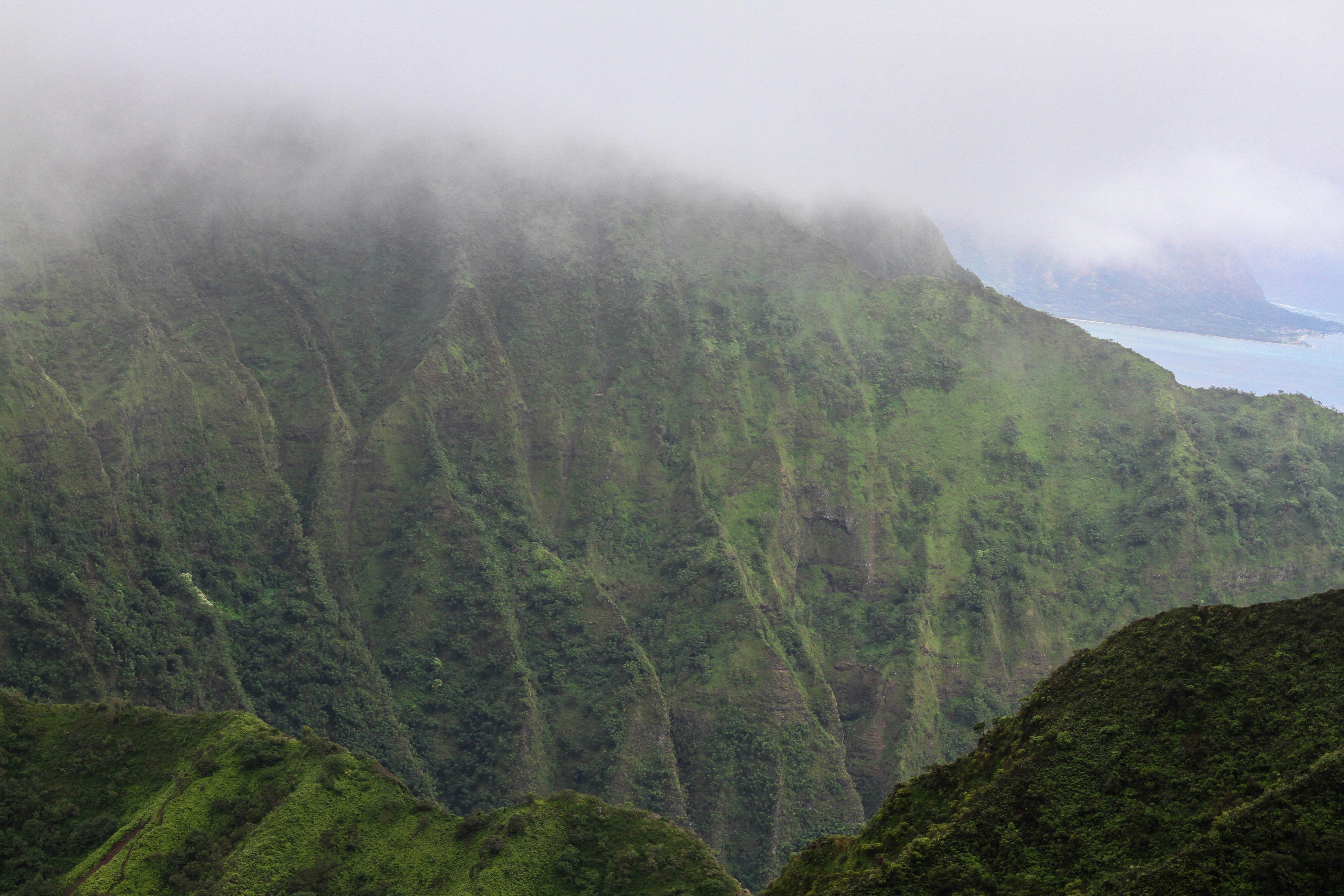 Lush green mountains interspersed with mist, revealing the intricate textures of the landscape. The coastline peeks through the clouds in the distance.