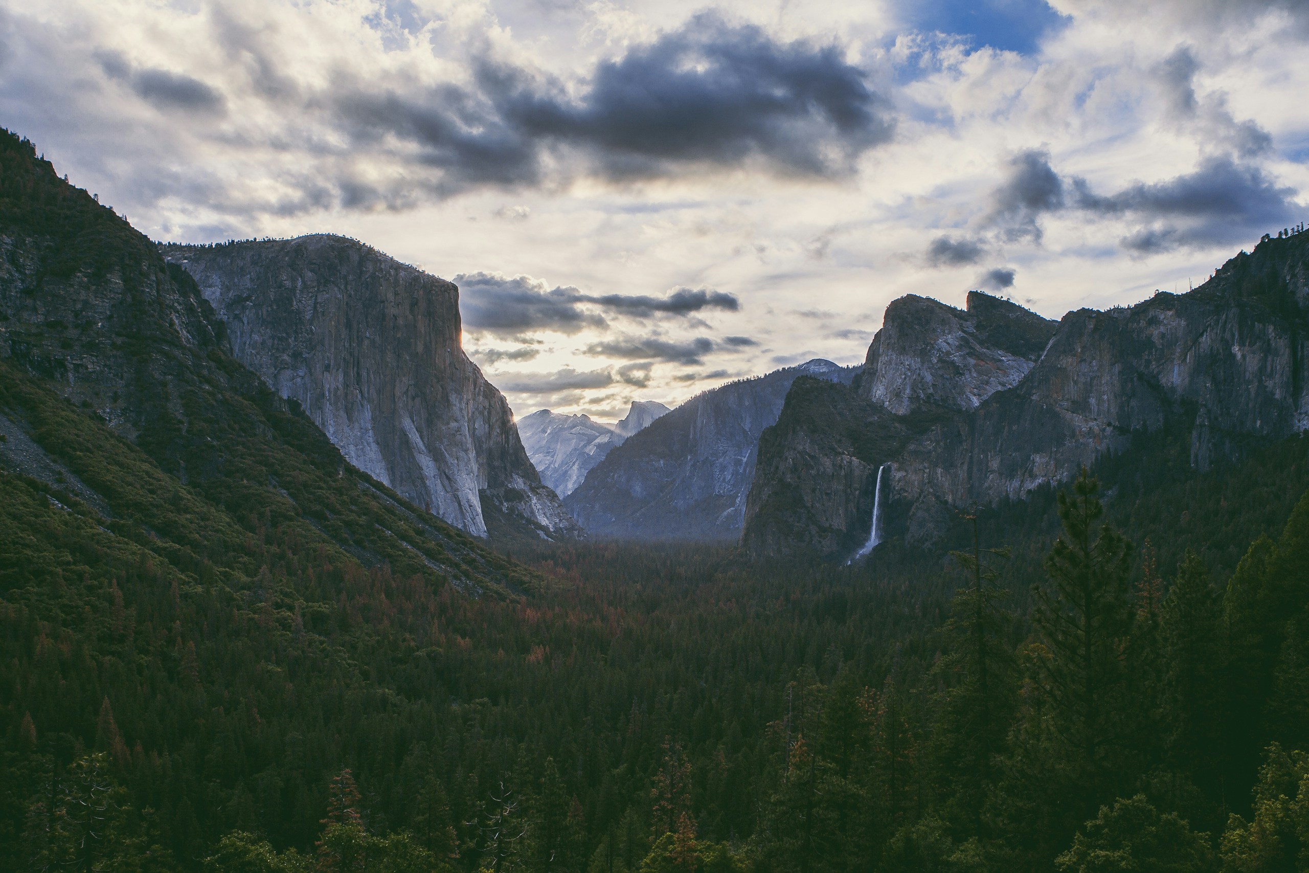 Mountainous landscape with dense forest and a distant waterfall under a cloud-filled sky.
